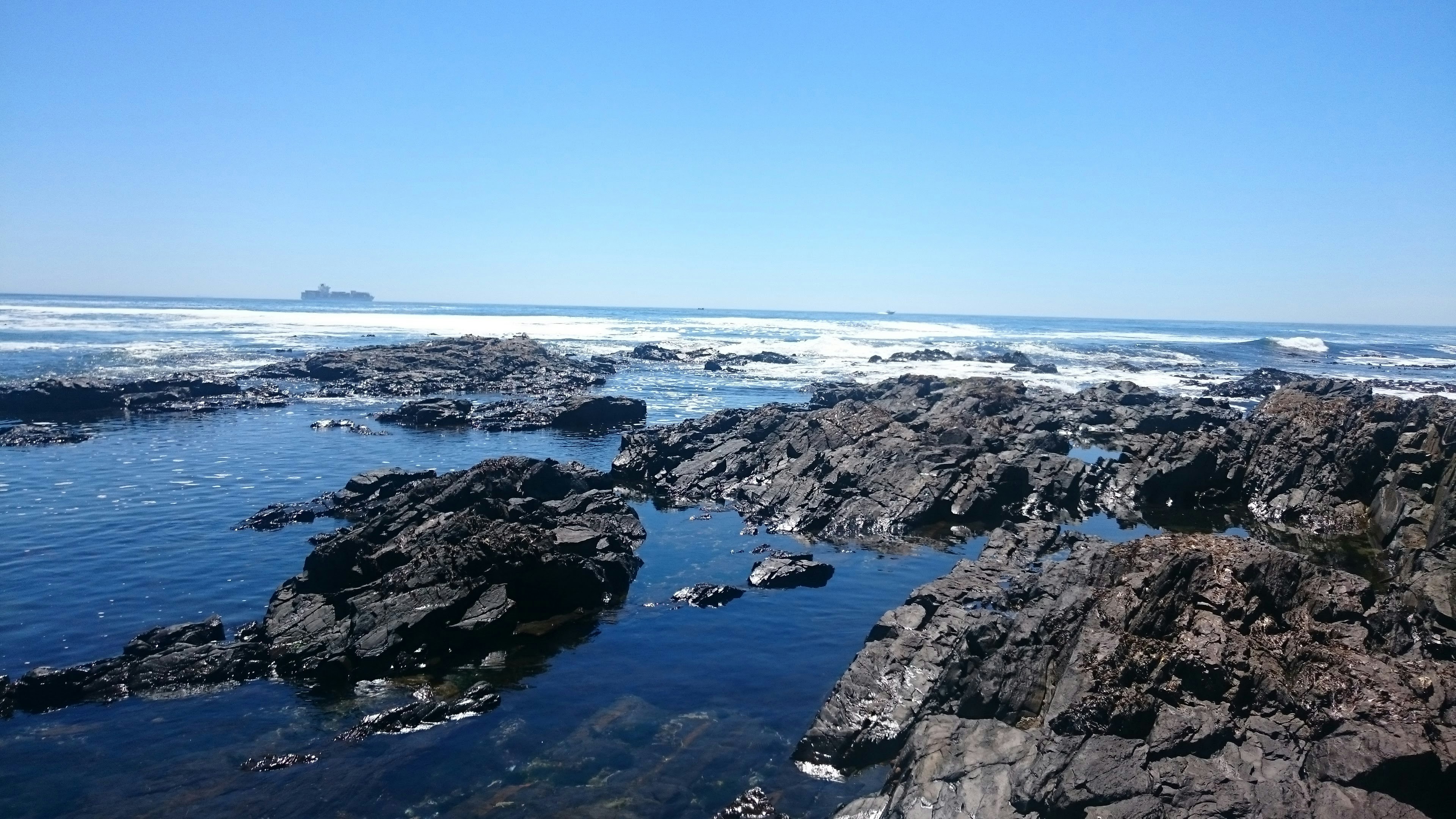 Rugged coastal rocks jutting into a clear blue sea, with gentle waves lapping against them under a bright sky.