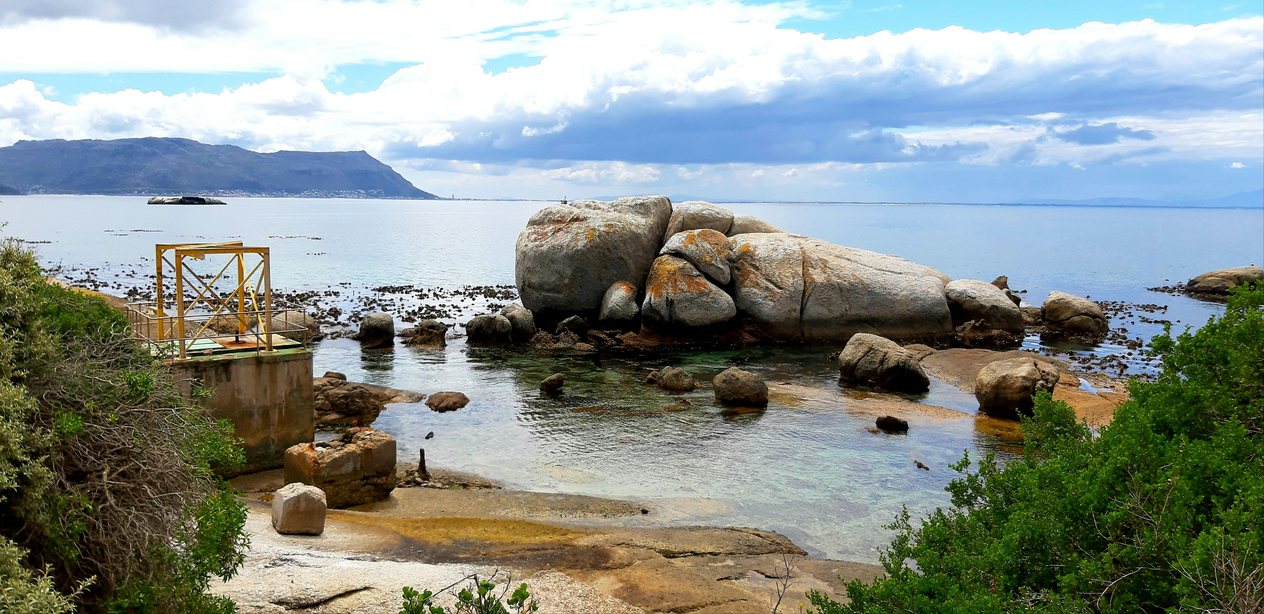 Rocky shoreline with a wooden structure, surrounded by calm waters and distant hills under a cloudy sky.