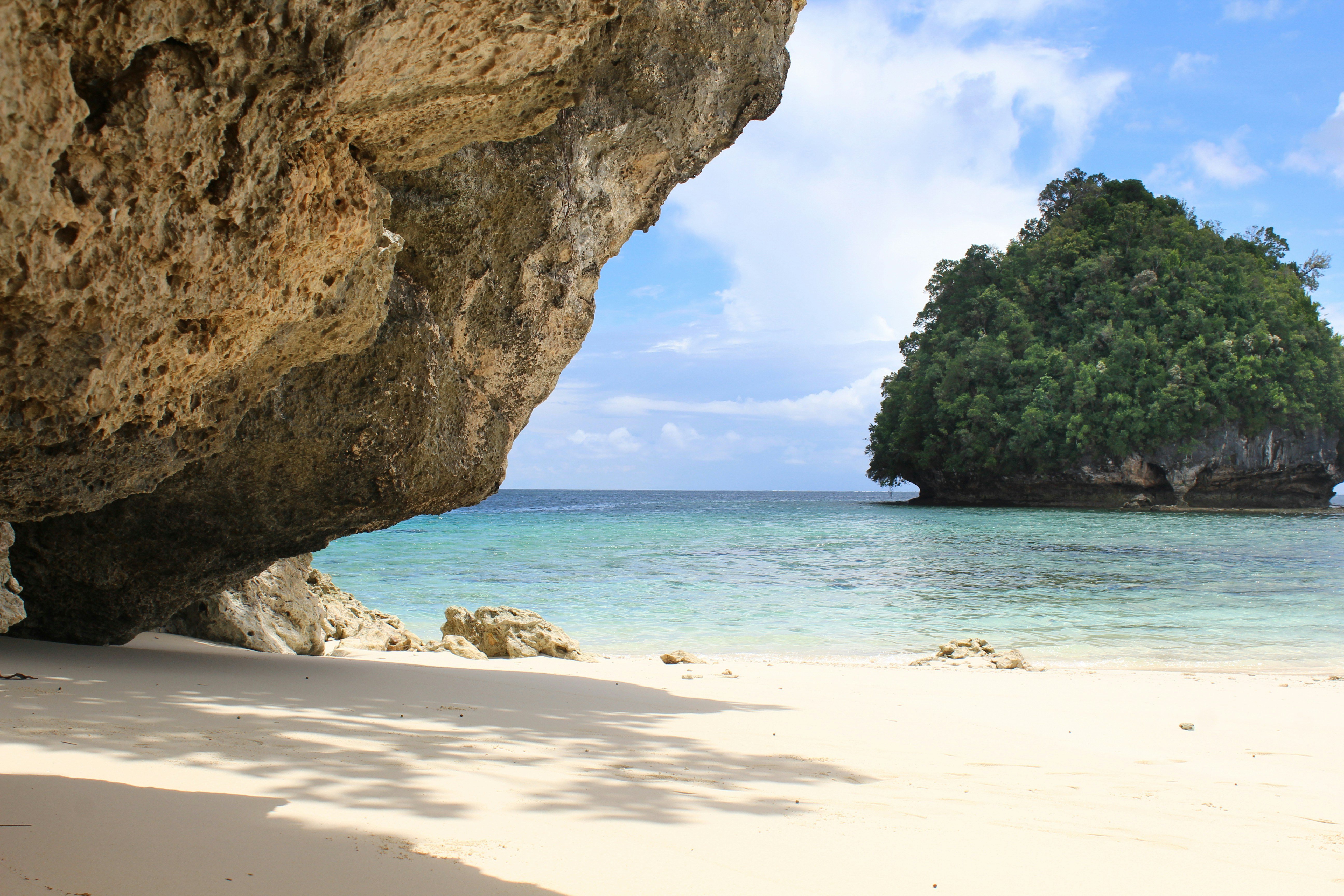 Rocky outcrop framing a tranquil beach scene with a lush island in the background. The clear blue waters invite exploration.