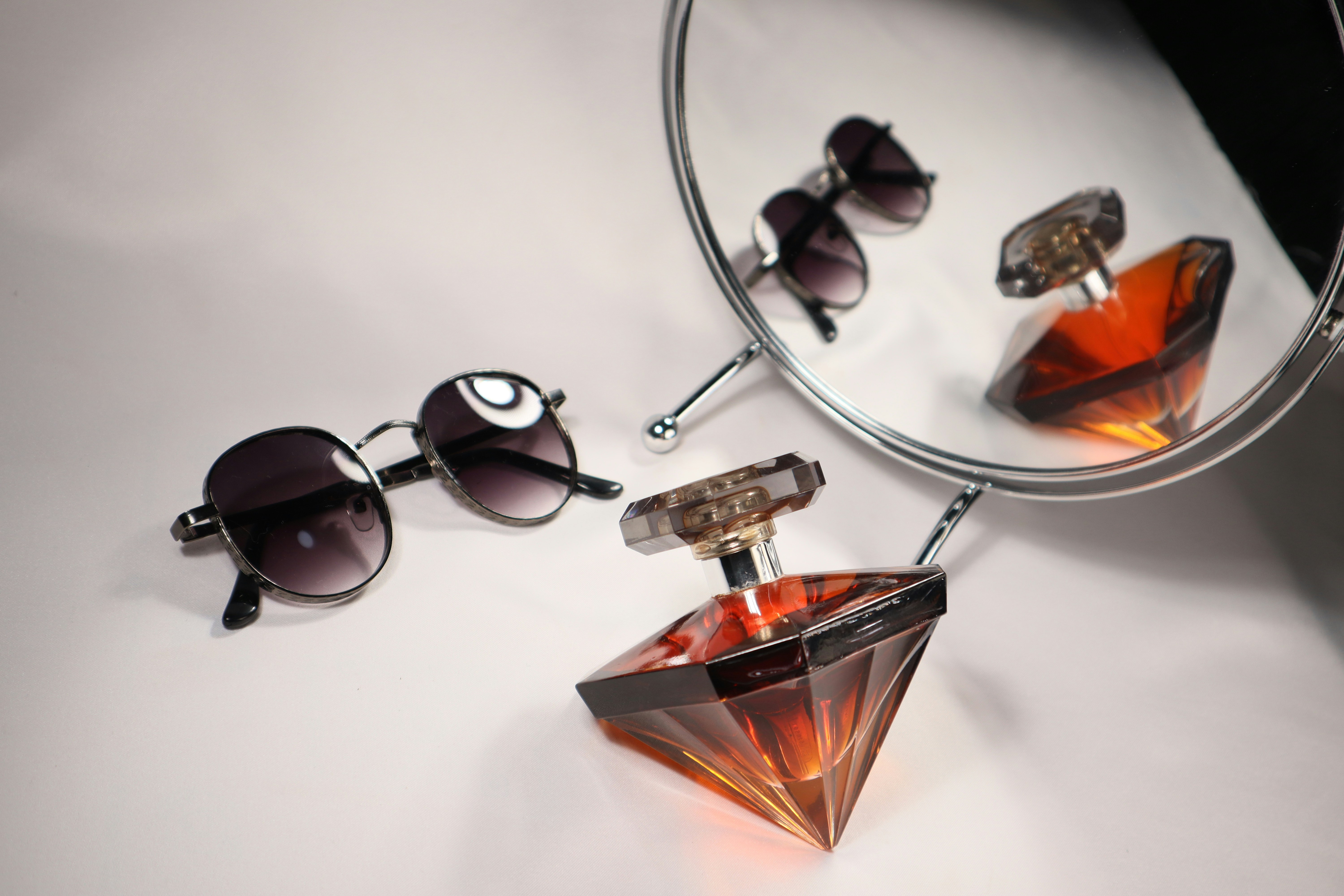 La Nuit Trésor Eau de Parfum by Lancôme is a fragrance that symbolizes the passion and elegance of an unforgettable night. It is an ode to femininity and love, designed to involve the wearer in a veil of sophistication and sensuality. https://fashionneedles.in/product/lancome-la-nuit-tresor-eau-de-parfum-for-women-75ml-tester/ | Sunglasses and perfume are reflected in a mirror.