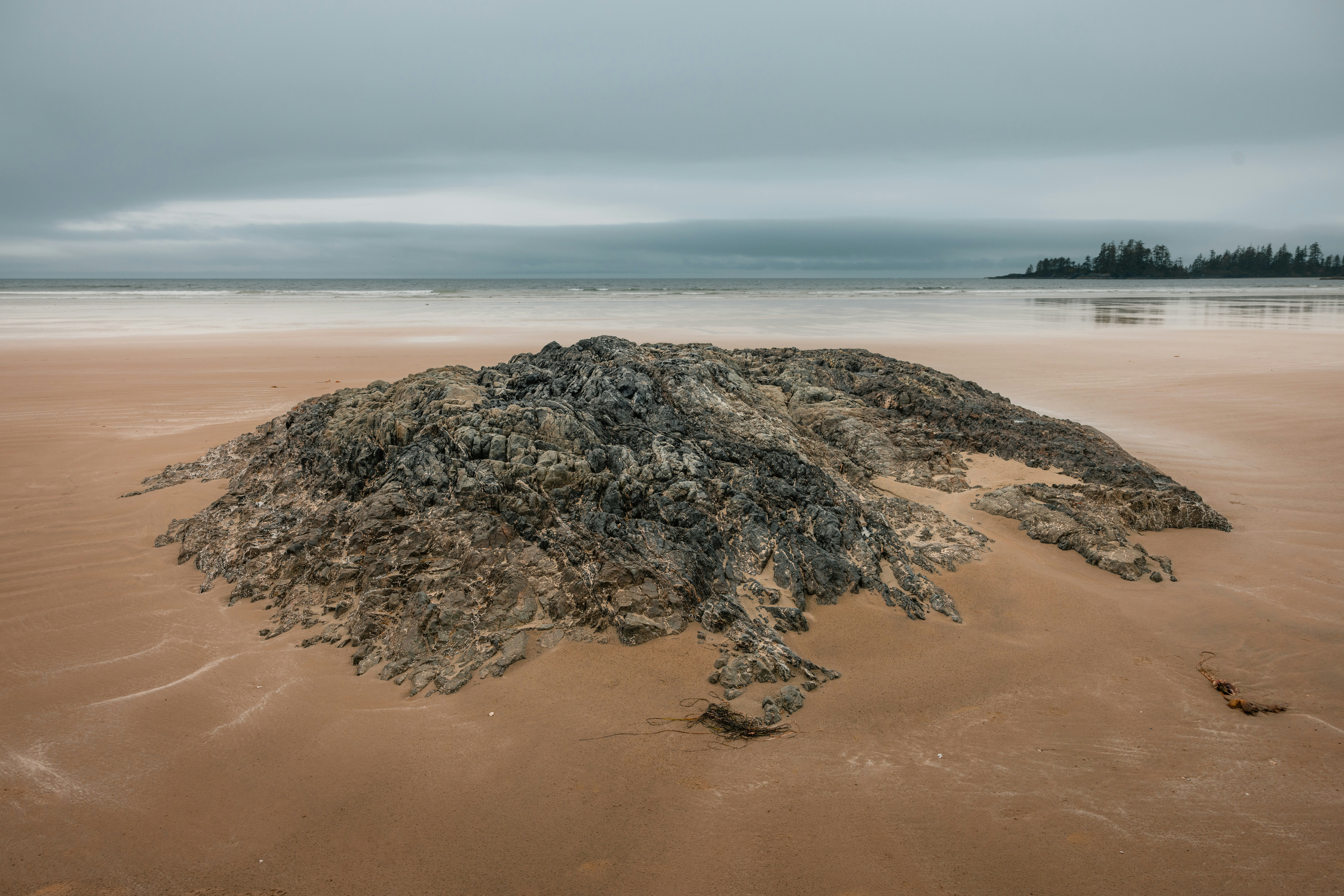 A rugged rock formation rises from the sandy beach, surrounded by a tranquil shoreline under an overcast sky.