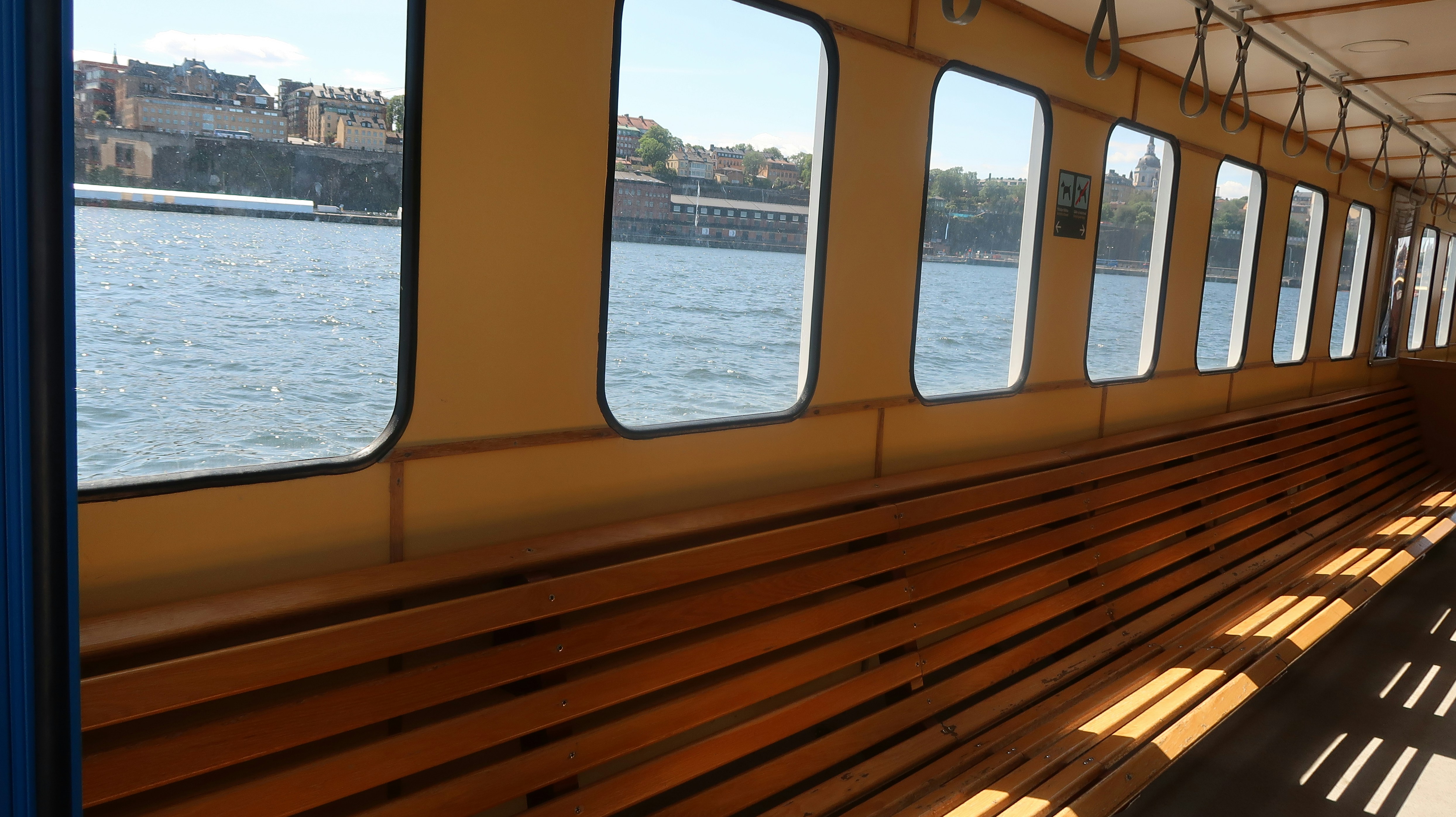 Interior view of a boat showcasing wooden benches and large windows overlooking the water. The sunlight casts patterns on the seating.