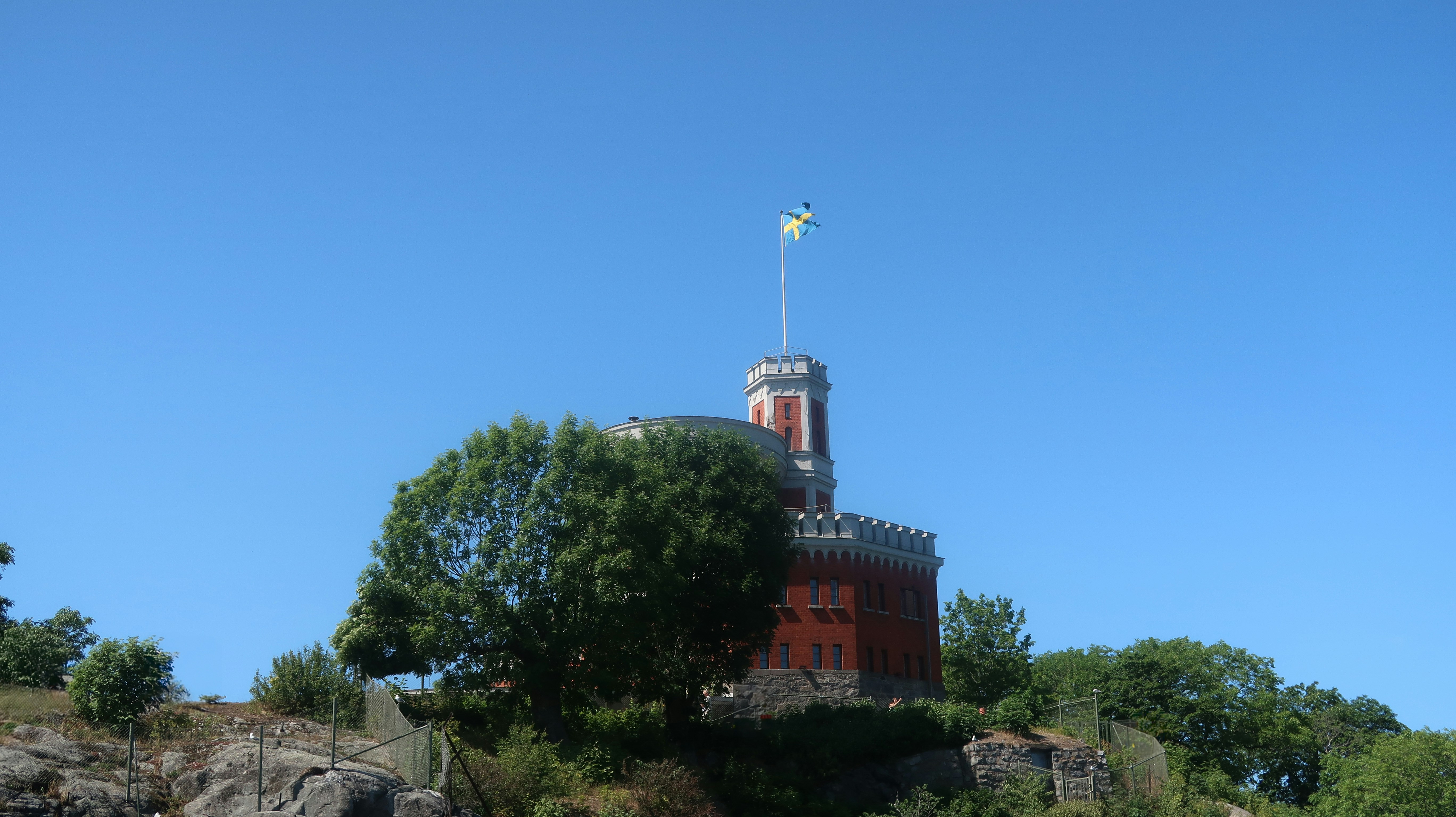 Historic red tower with a flag, surrounded by lush greenery and rocky terrain.