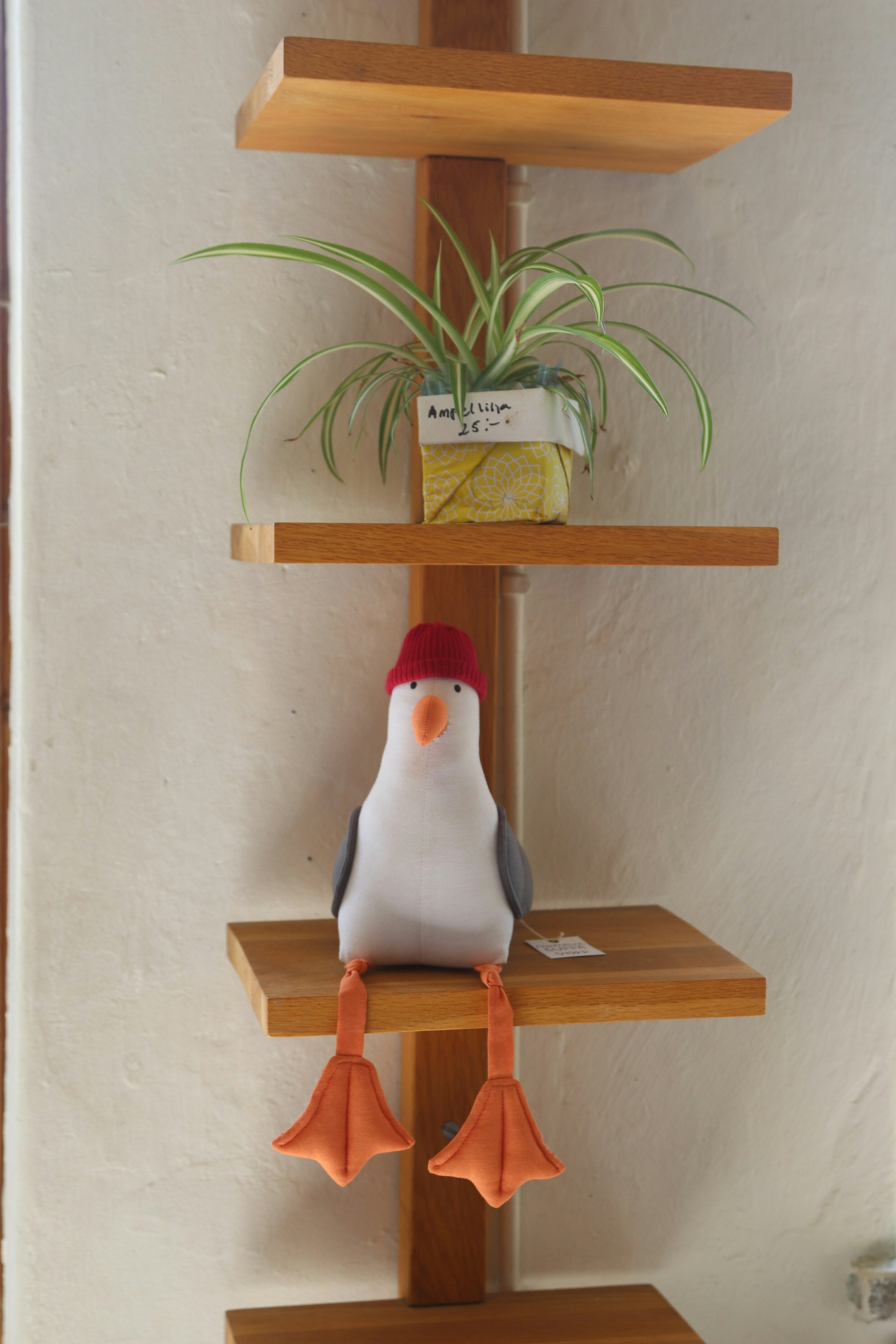 A whimsical plush seagull adorned with a red hat and orange flippers sits on a wooden shelf, accompanied by a small potted plant.
