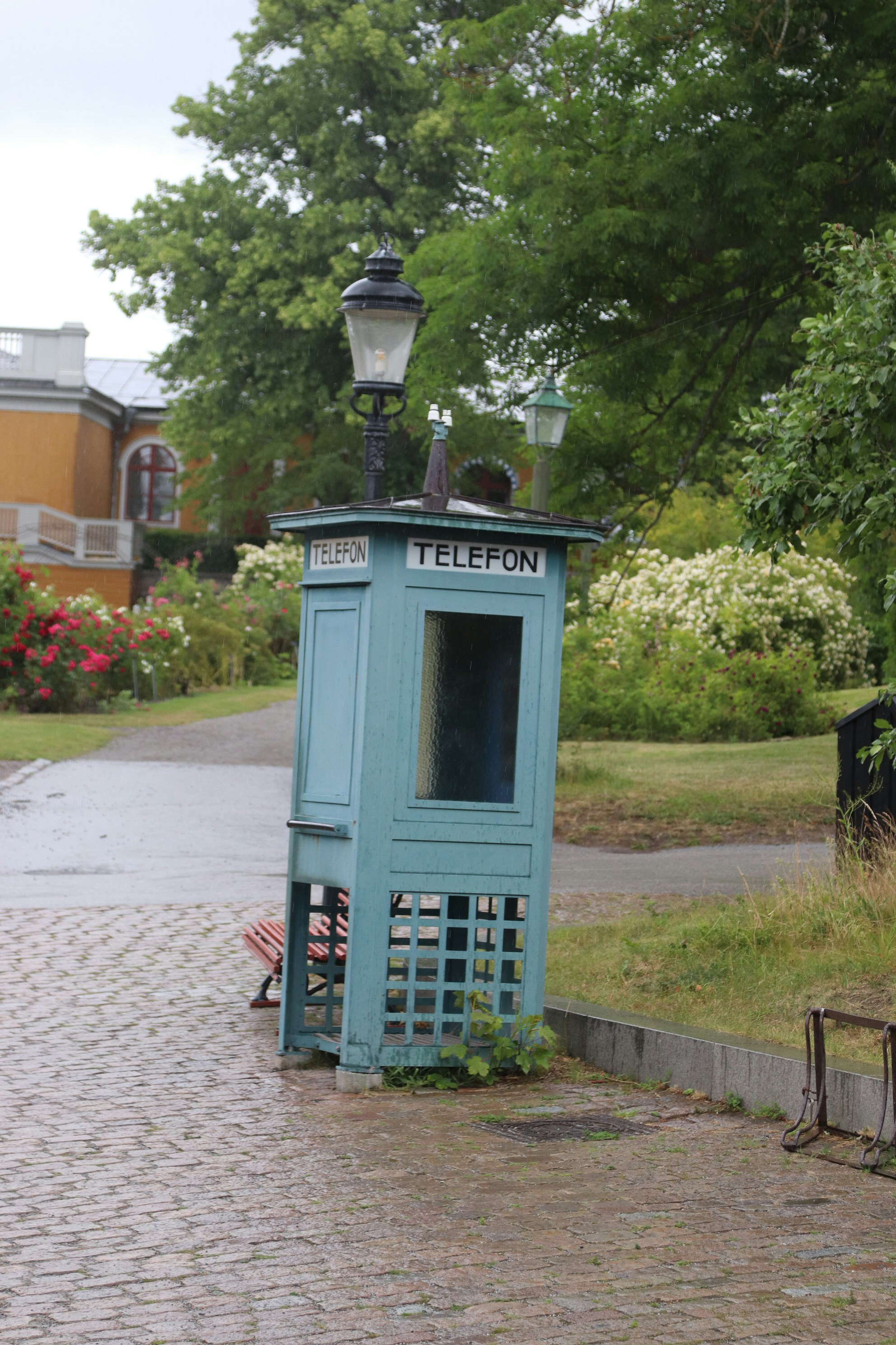 A weathered blue telephone booth stands amidst lush greenery and blooming flowers, evoking nostalgia in a serene park setting.