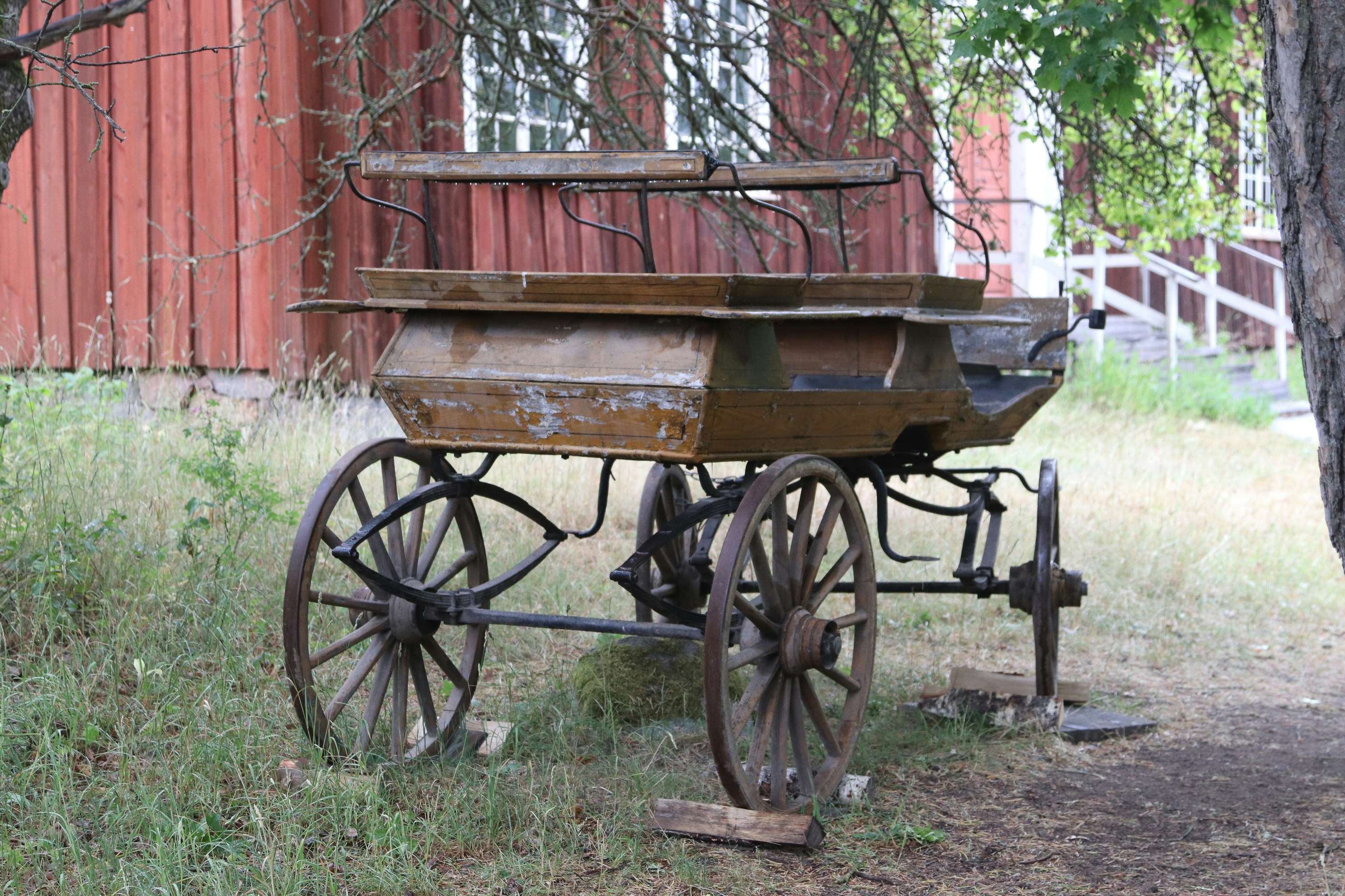 Weathered wooden cart resting on overgrown grass beside a rustic red barn. The scene evokes nostalgia and a sense of history.