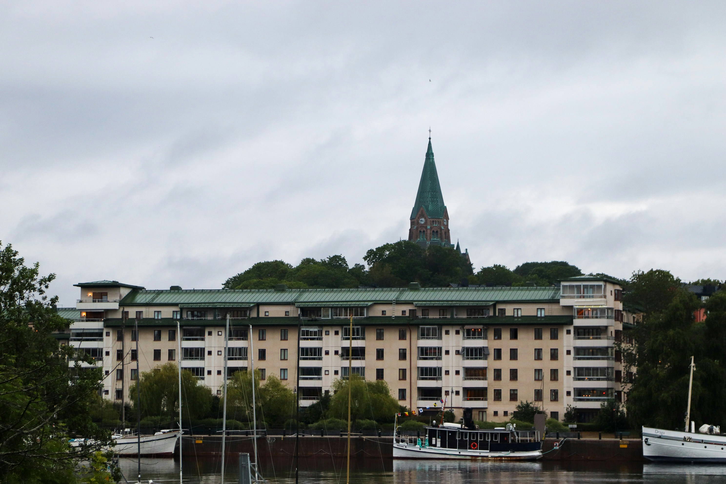 Historic church steeple rises above contemporary waterfront apartments, framed by lush greenery and boats at dock.