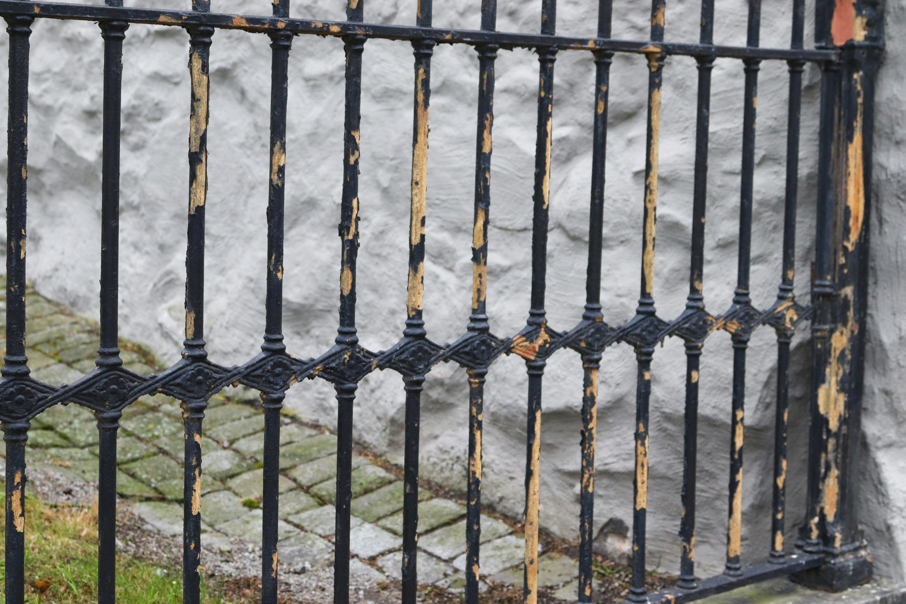 Weathered black iron gate with peeling paint, set against a textured gray wall and cobblestone path.