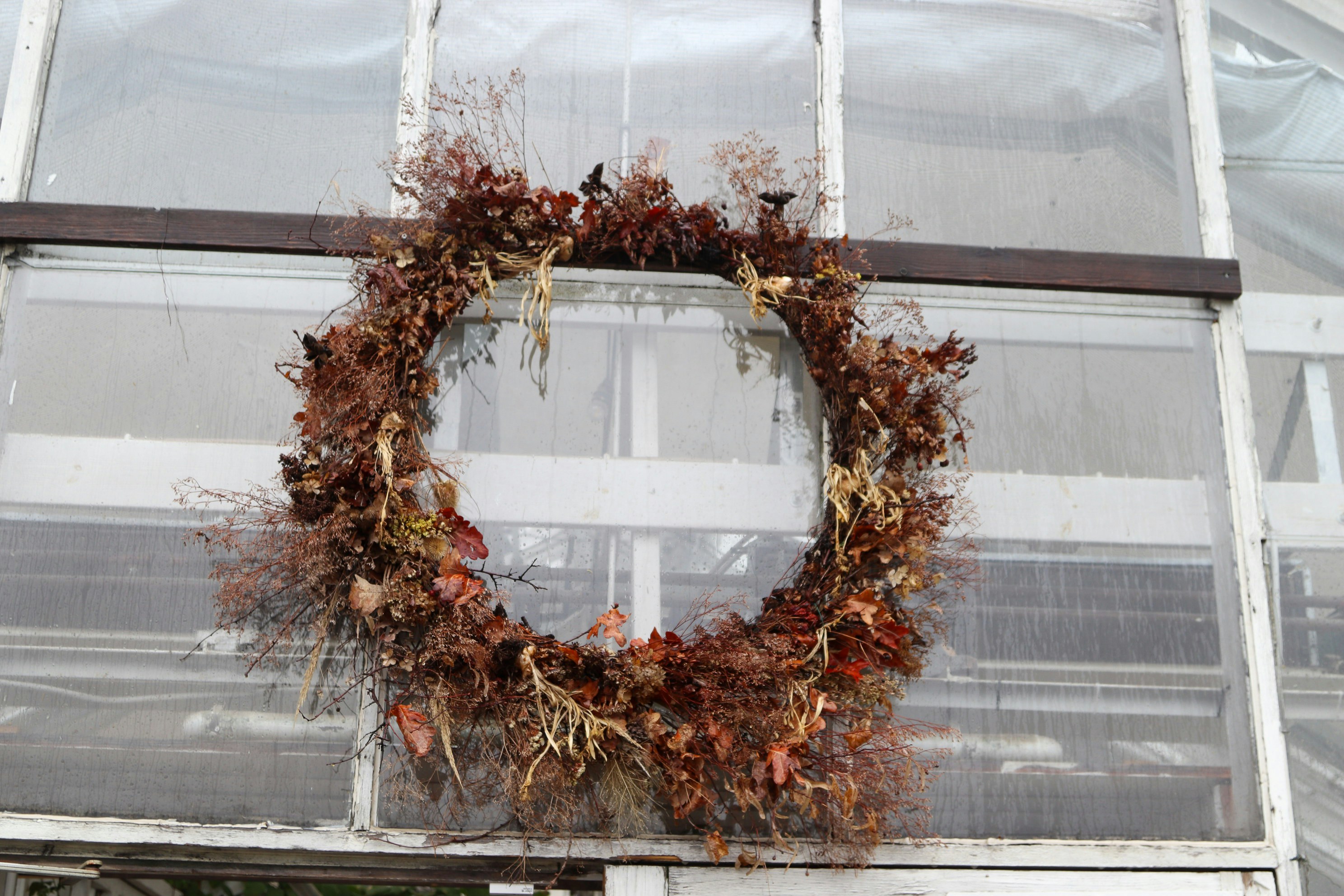 Dried floral wreath adorned with autumn leaves, suspended against a backdrop of glass and wood. A testament to seasonal change.