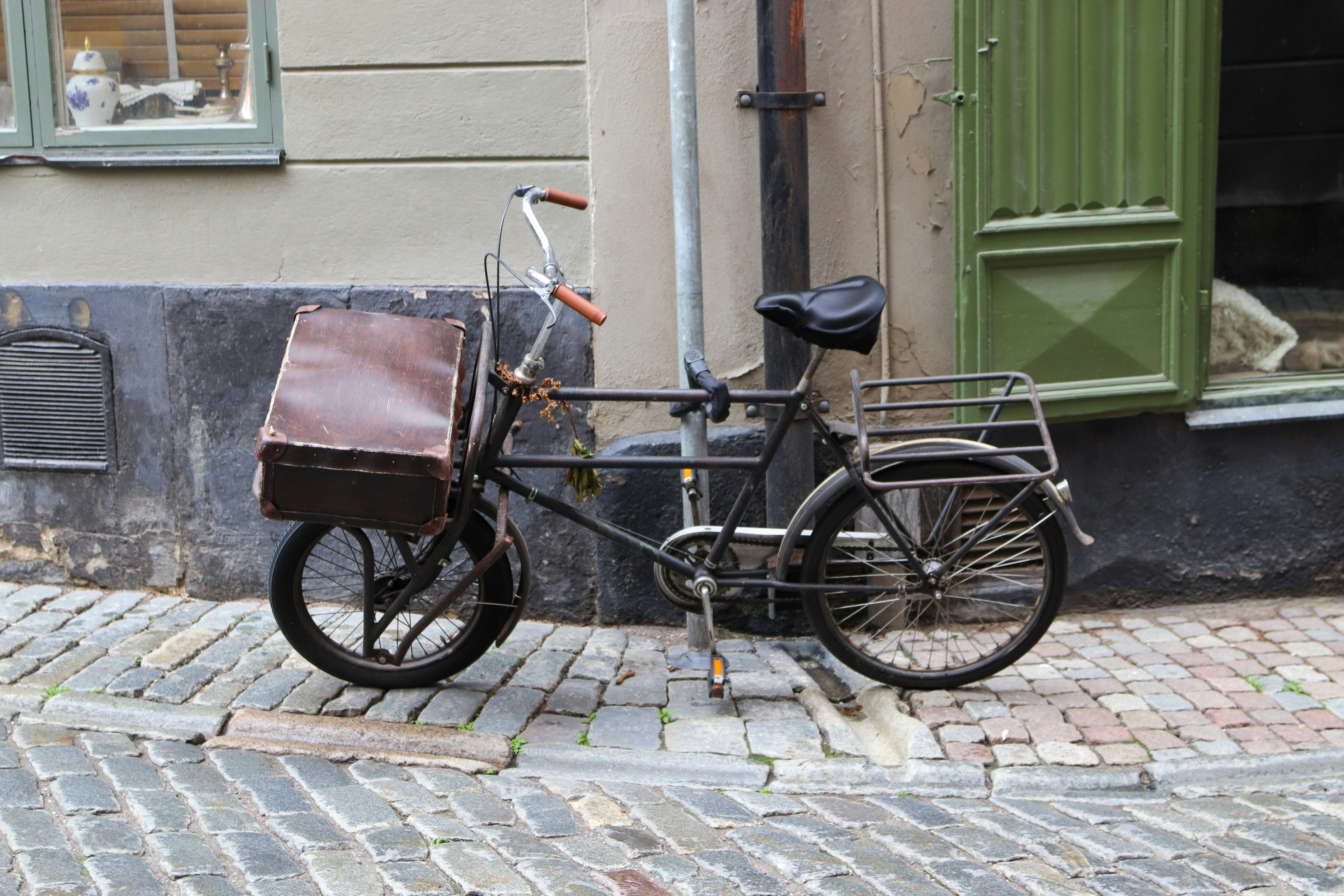 An old bicycle with a leather box rests against a cobblestone street, framed by a quaint building and a green window. The scene evokes a sense of nostalgia and urban charm.