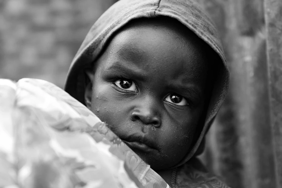 Black-and-white close-up of a young child in a hooded sweatshirt, looking directly at the camera with a serious expression. A patterned cloth fills the lower left foreground.