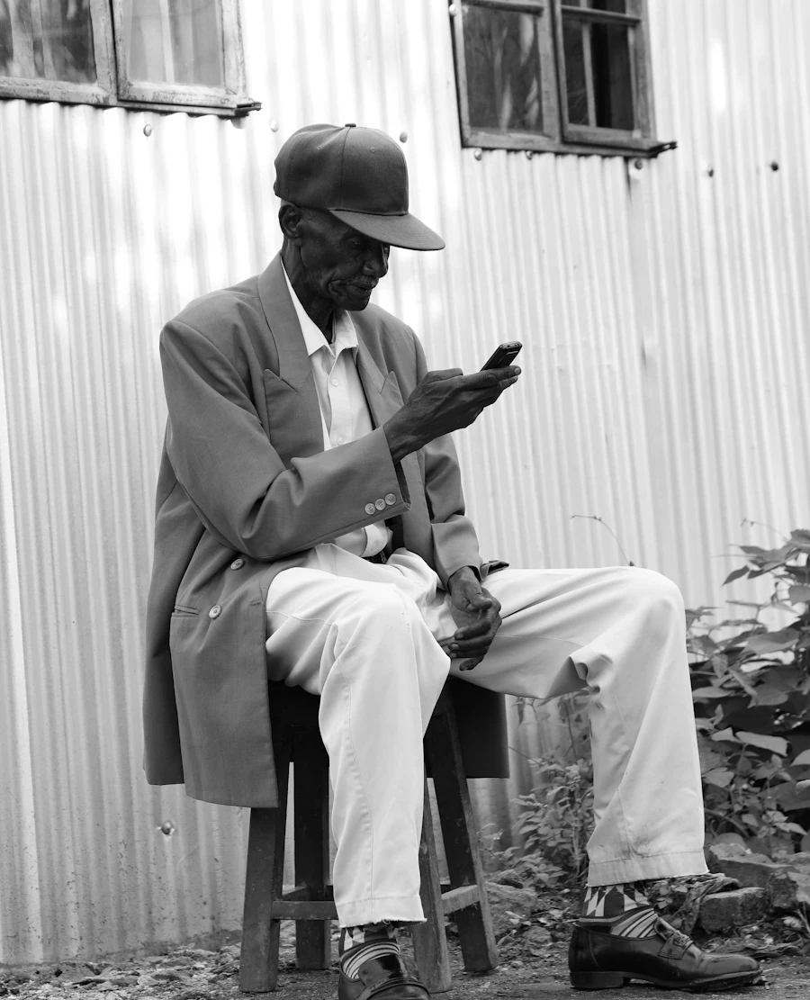 An elderly man in a blazer and cap sits on a stool outdoors, looking down at a phone in his hand. Corrugated metal siding and windows form a stark black-and-white background.