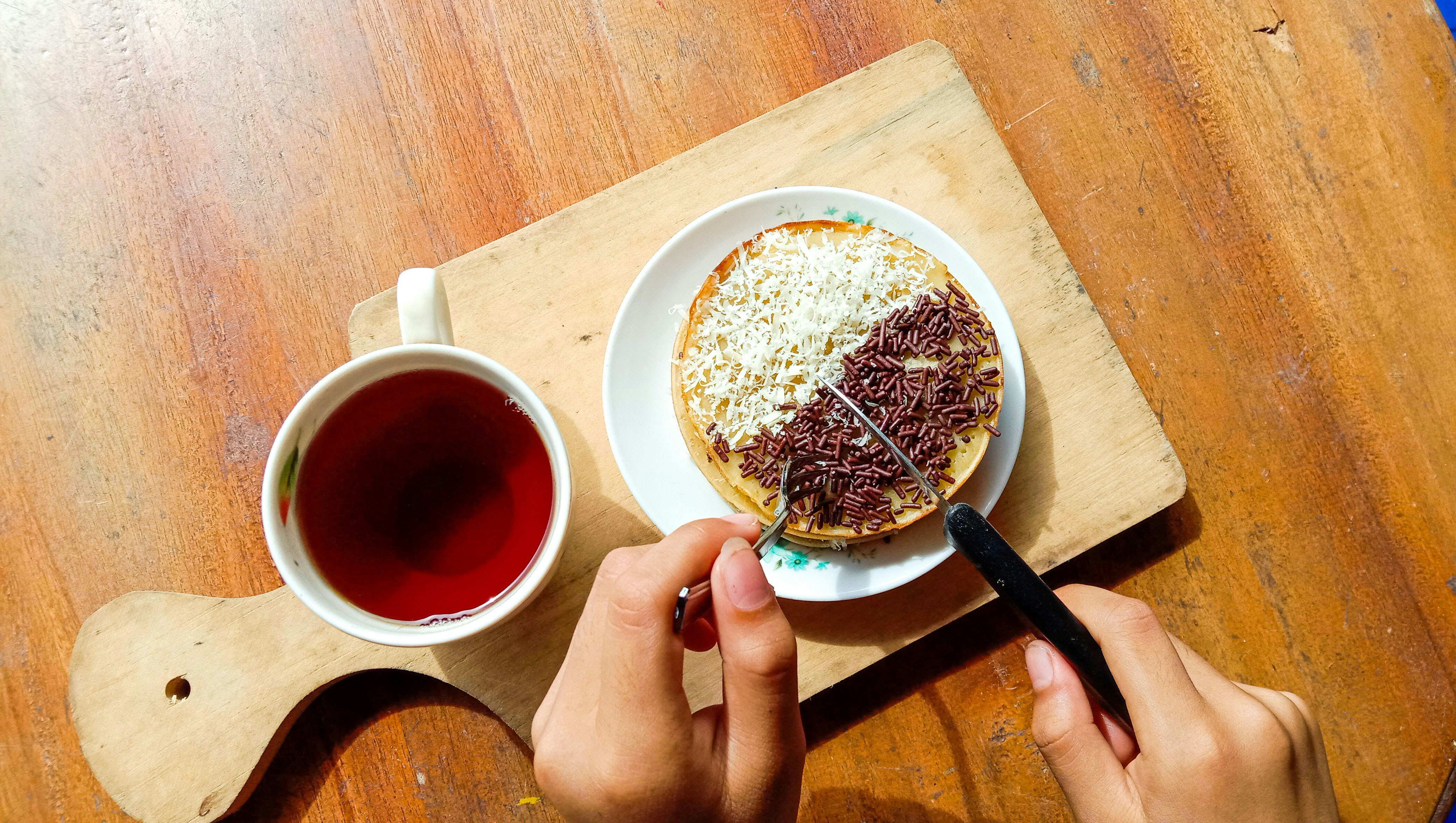 A mini photo of a martabak with a chocolate topping and grated cheese found a cup is hot, a knife, a fork and cutlery | Someone is cutting a delicious dessert and drinking tea.