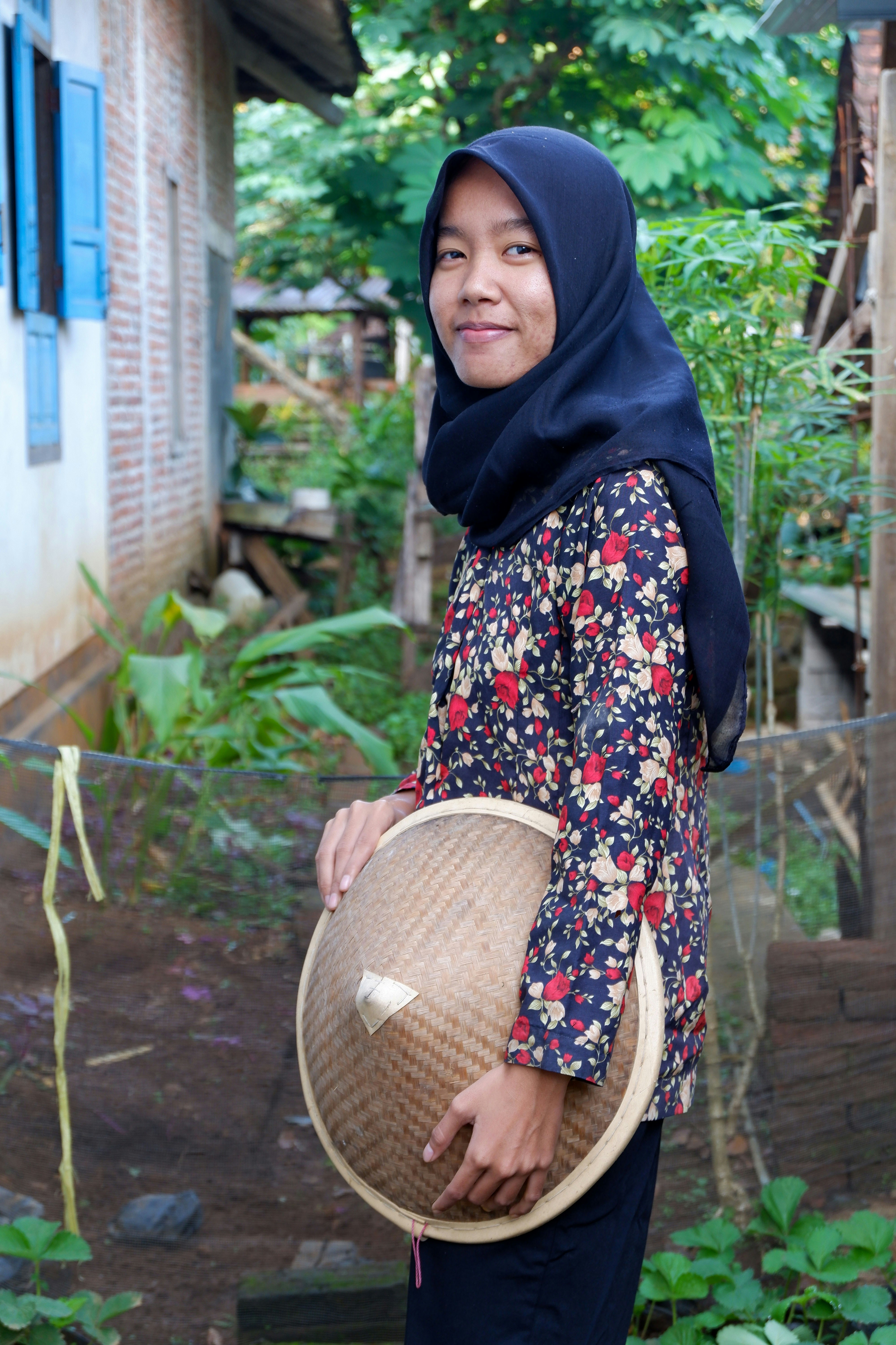 December 18, 2019, photo of a woman dressed in a typical Indonesian batik planting strawberry plants during the day, Wadaslintang, Wonosobo, Indonesia | A woman wearing a hijab holds a woven hat.