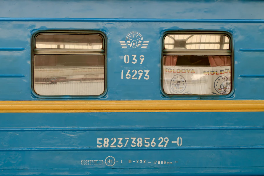 Blue passenger rail carriage side showing two rectangular windows, Moldova emblem and curtain visible through the right window and white stenciled fleet number on the lower panel.