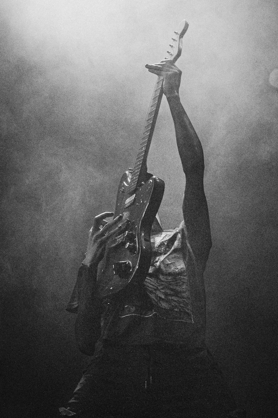A guitarist silhouetted in heavy smoke holds the electric guitar overhead under dramatic concert lighting.