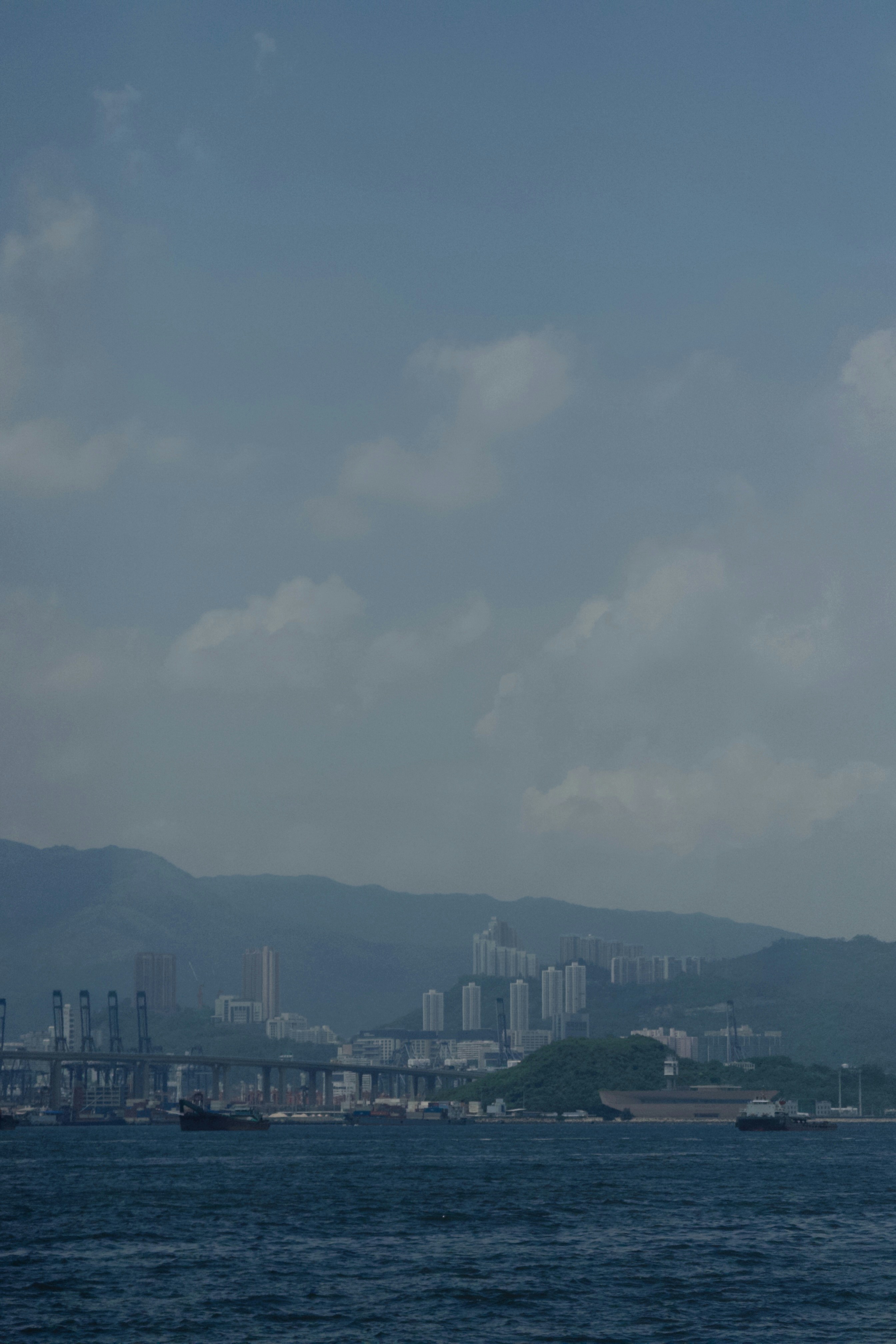 A tranquil harbor scene featuring distant city skyscrapers and lush hills under a soft, cloudy sky.