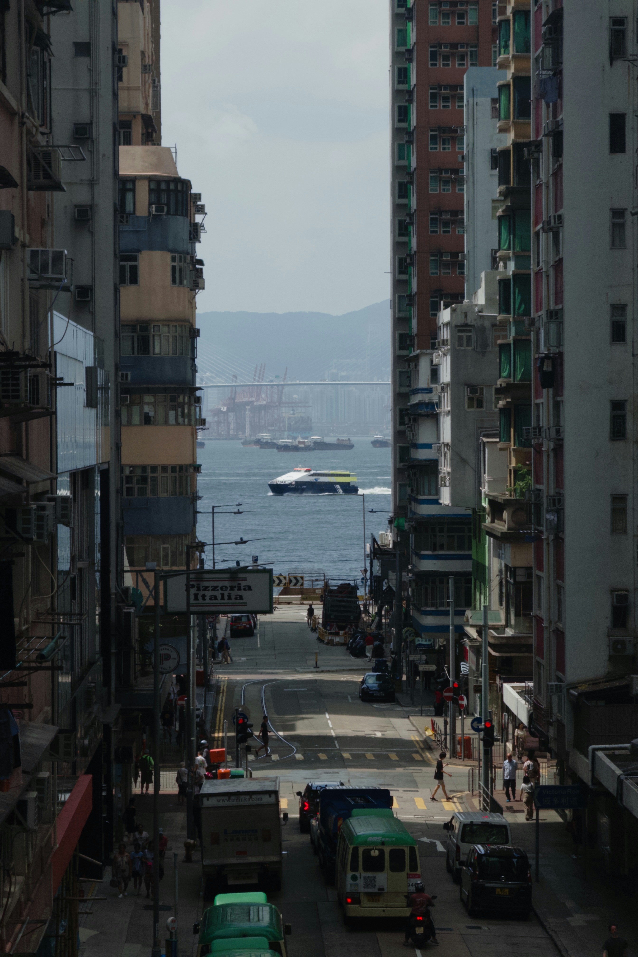 View down a bustling city street framed by high-rise buildings, showcasing a ferry navigating the harbor in the background.