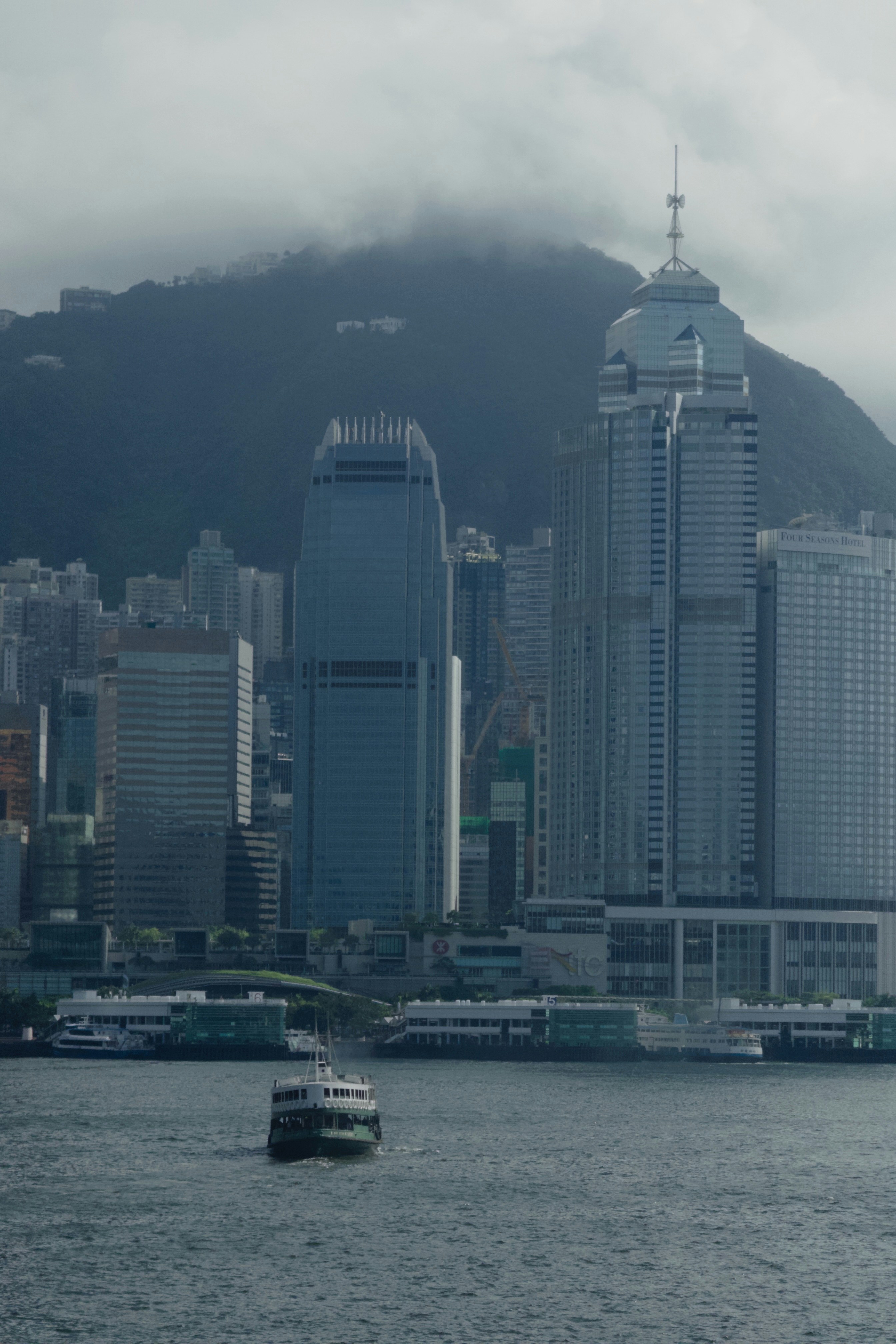 A ferry glides through the harbor, framed by towering skyscrapers and a misty mountain backdrop. The interplay of urban architecture and natural elements creates a striking contrast.