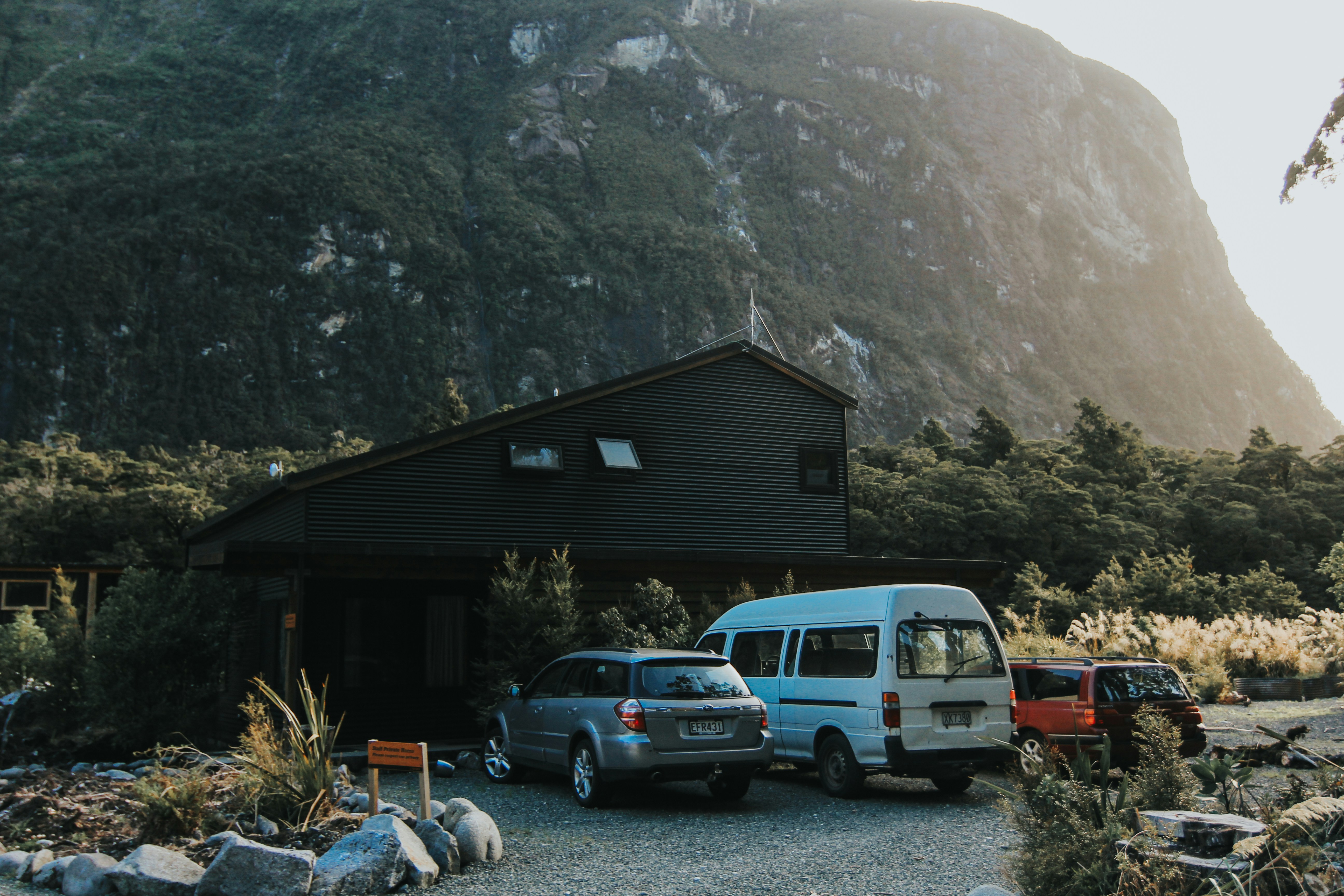 Modern house nestled in a mountainous landscape, surrounded by lush greenery and parked vehicles.