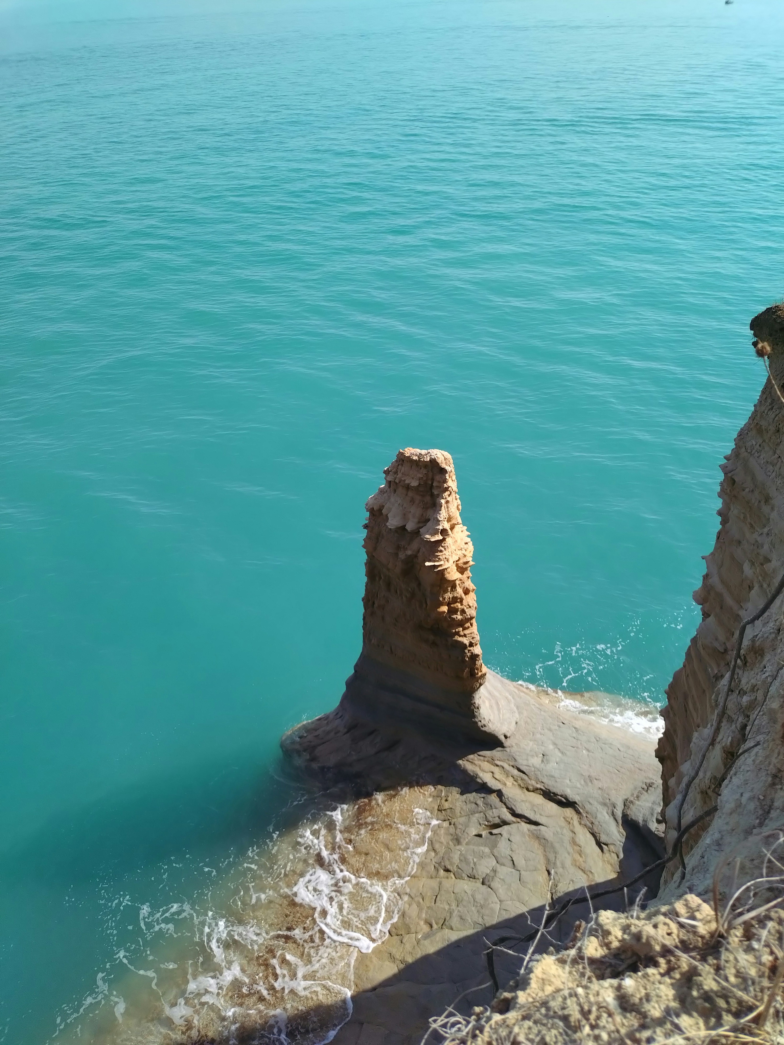 Lone rock formation stands defiantly against the turquoise sea, showcasing nature's sculpting power. Waves gently lap at the base, hinting at the relentless forces of erosion.
