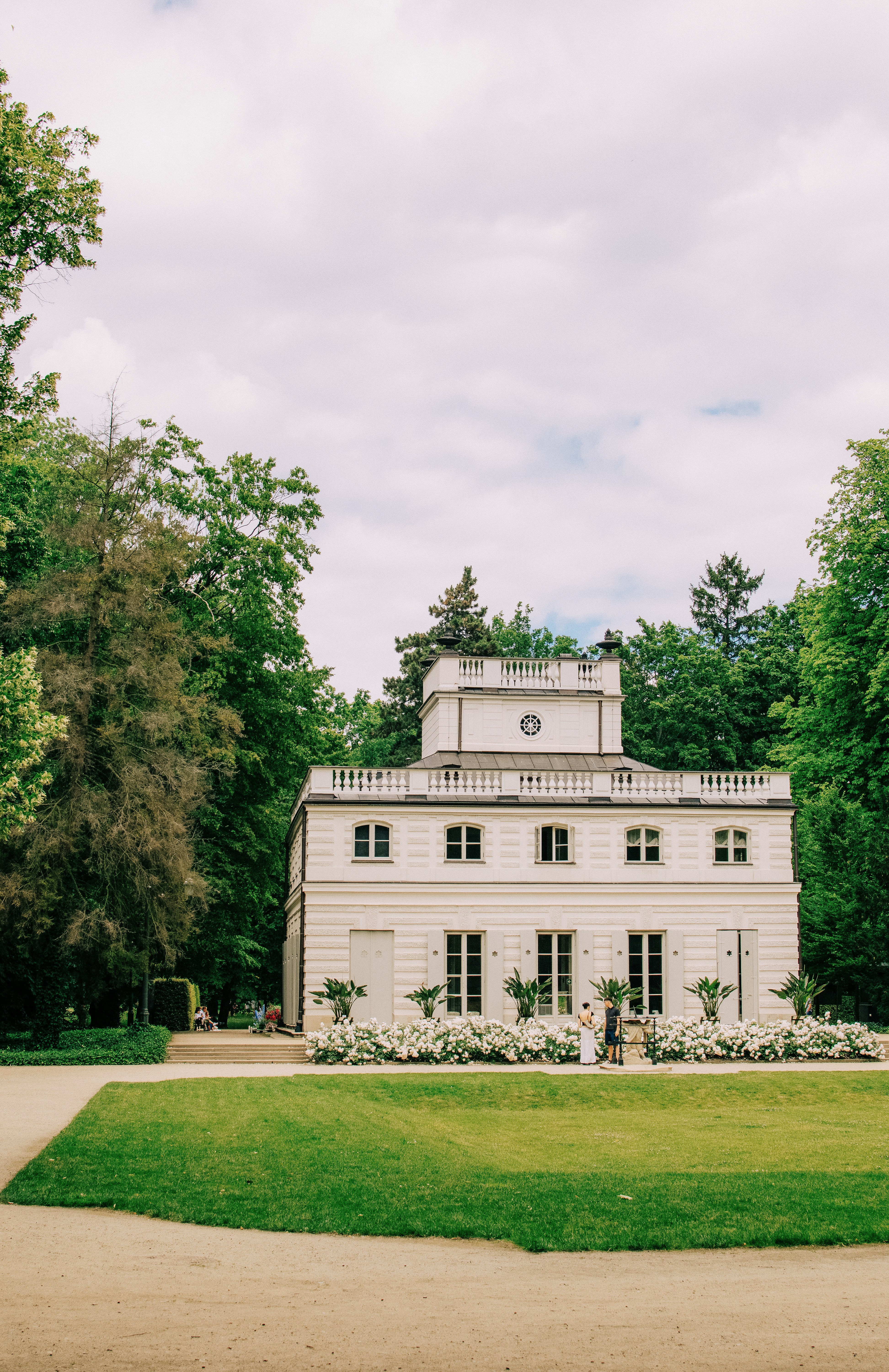 Charming white villa surrounded by lush greenery and blooming flowers, set against a cloudy sky.