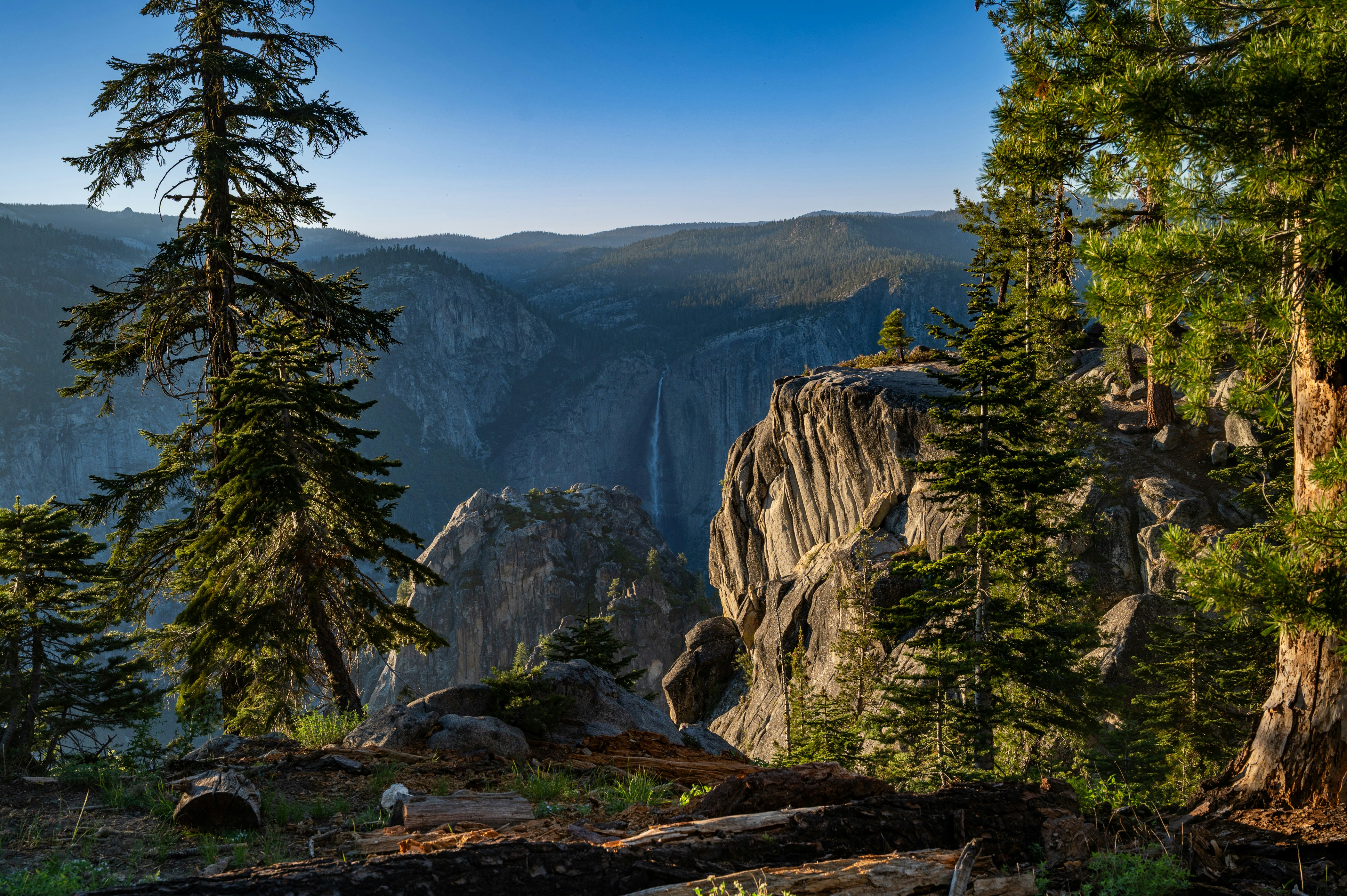 Majestic cliffs framed by towering pines, revealing a distant waterfall cascading into the valley below.