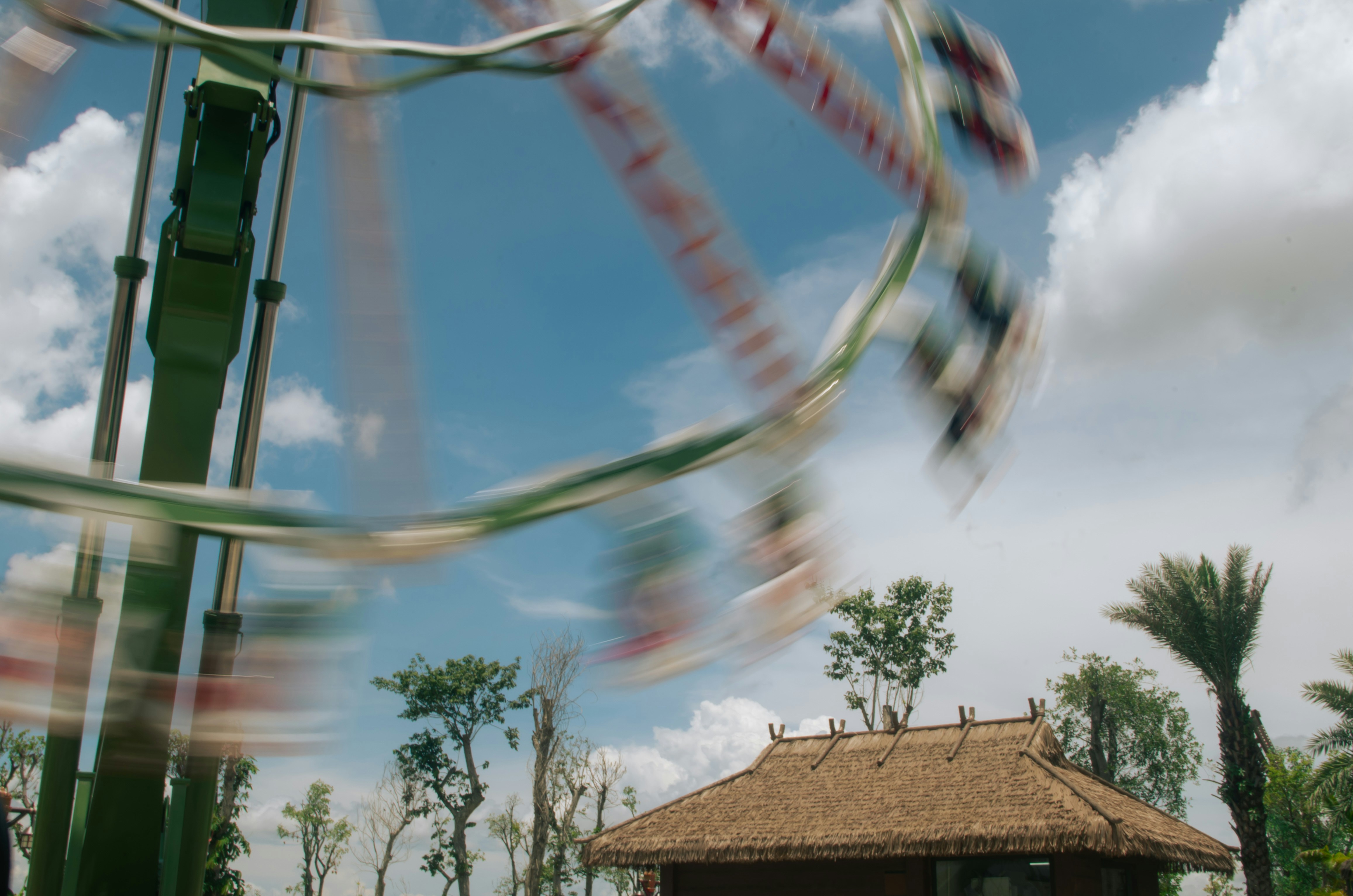 A vibrant ferris wheel spins against a backdrop of fluffy clouds and lush greenery, evoking a sense of fun and excitement. The traditional hut adds a cultural touch to the lively scene.