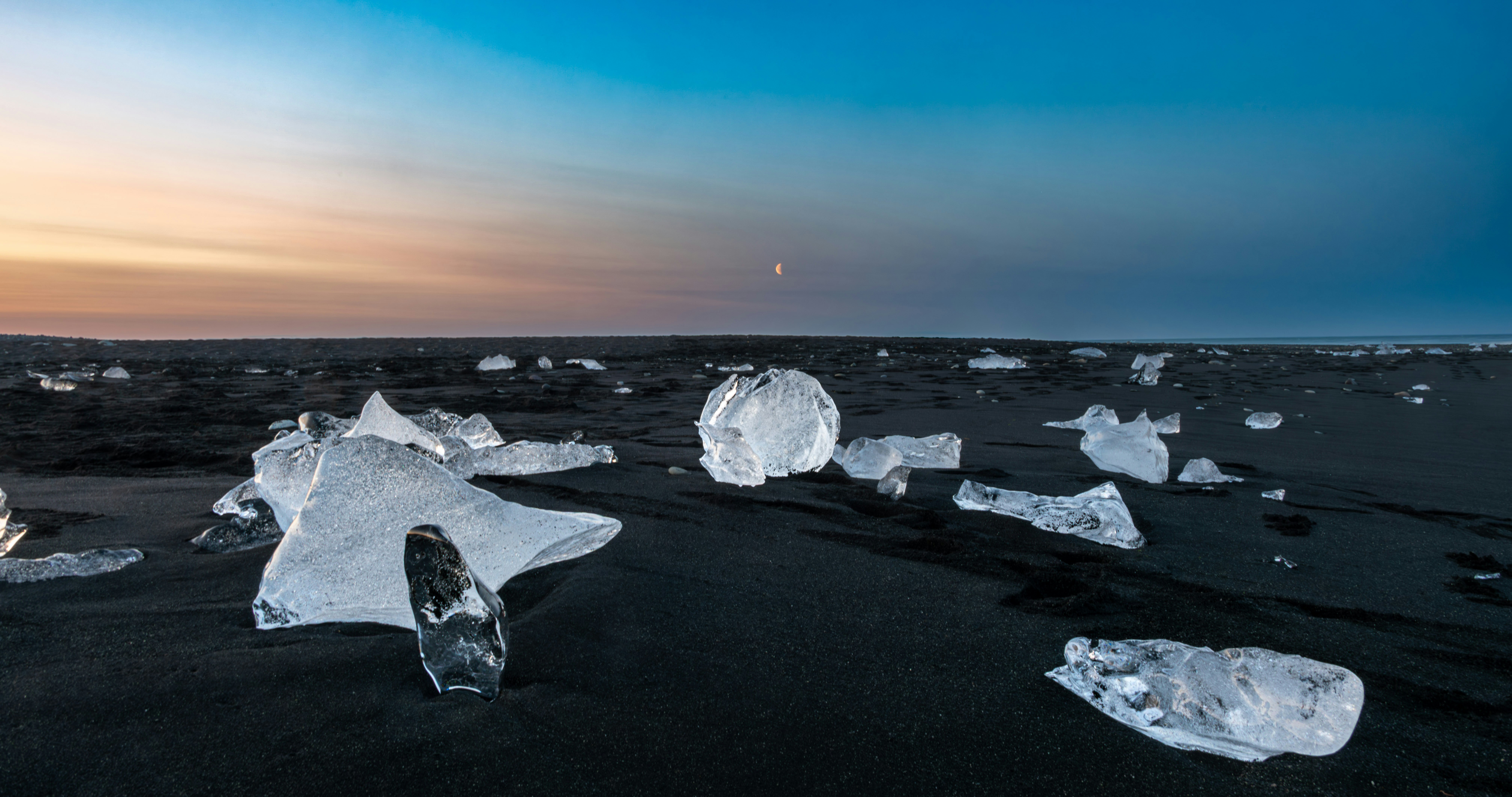 Translucent ice formations scattered across a black sandy beach at twilight, with a faint celestial body visible in the sky.