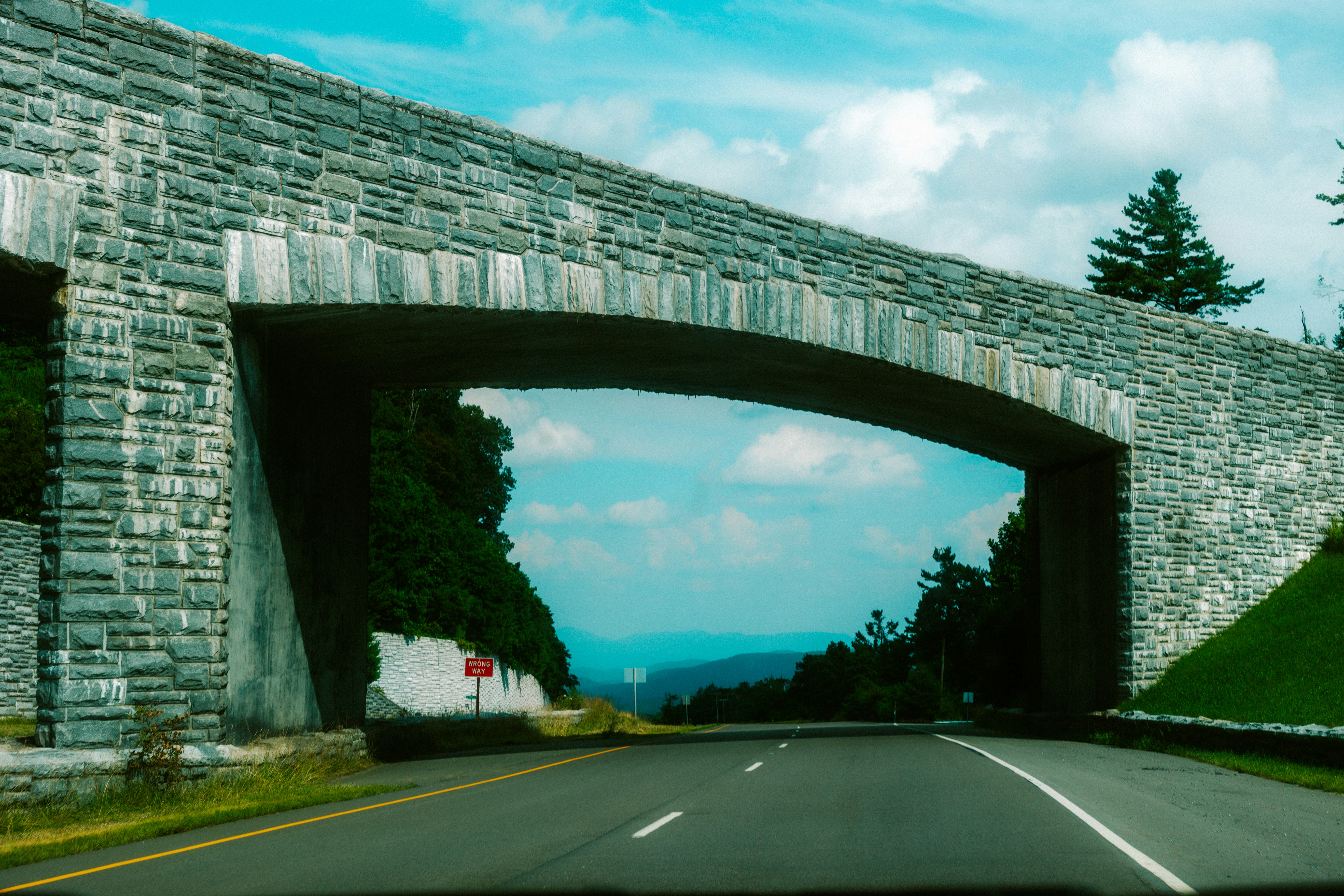 A stone arch bridge spans a highway, framed by lush greenery and distant mountains under a cloudy sky.