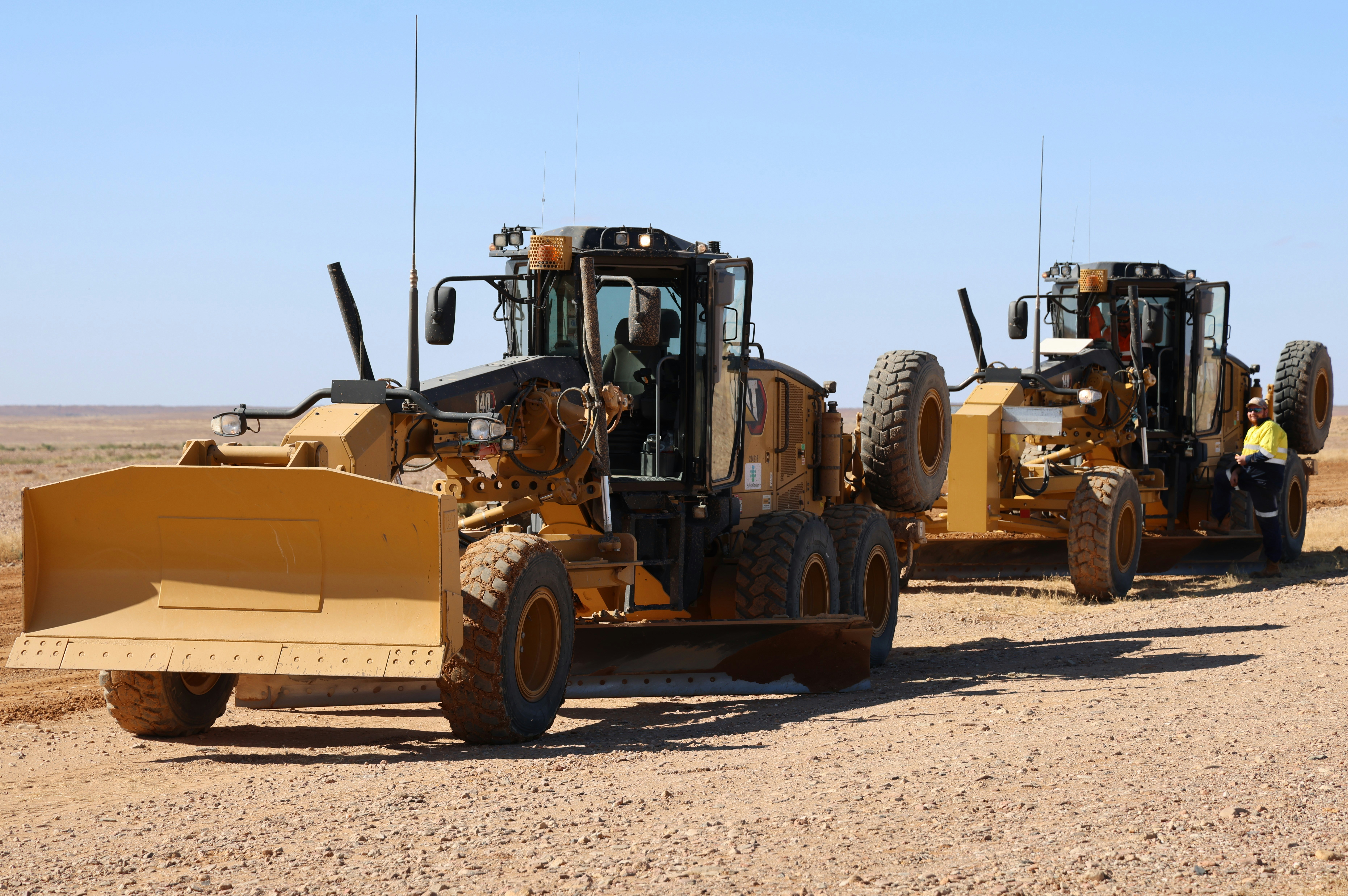 Twin graders resting on red dirt—silent giants poised for another day shaping the outback roads of South Australia. on the Oodnadatta Track in just north of Marree. | Two heavy-duty construction vehicles are on the ground.