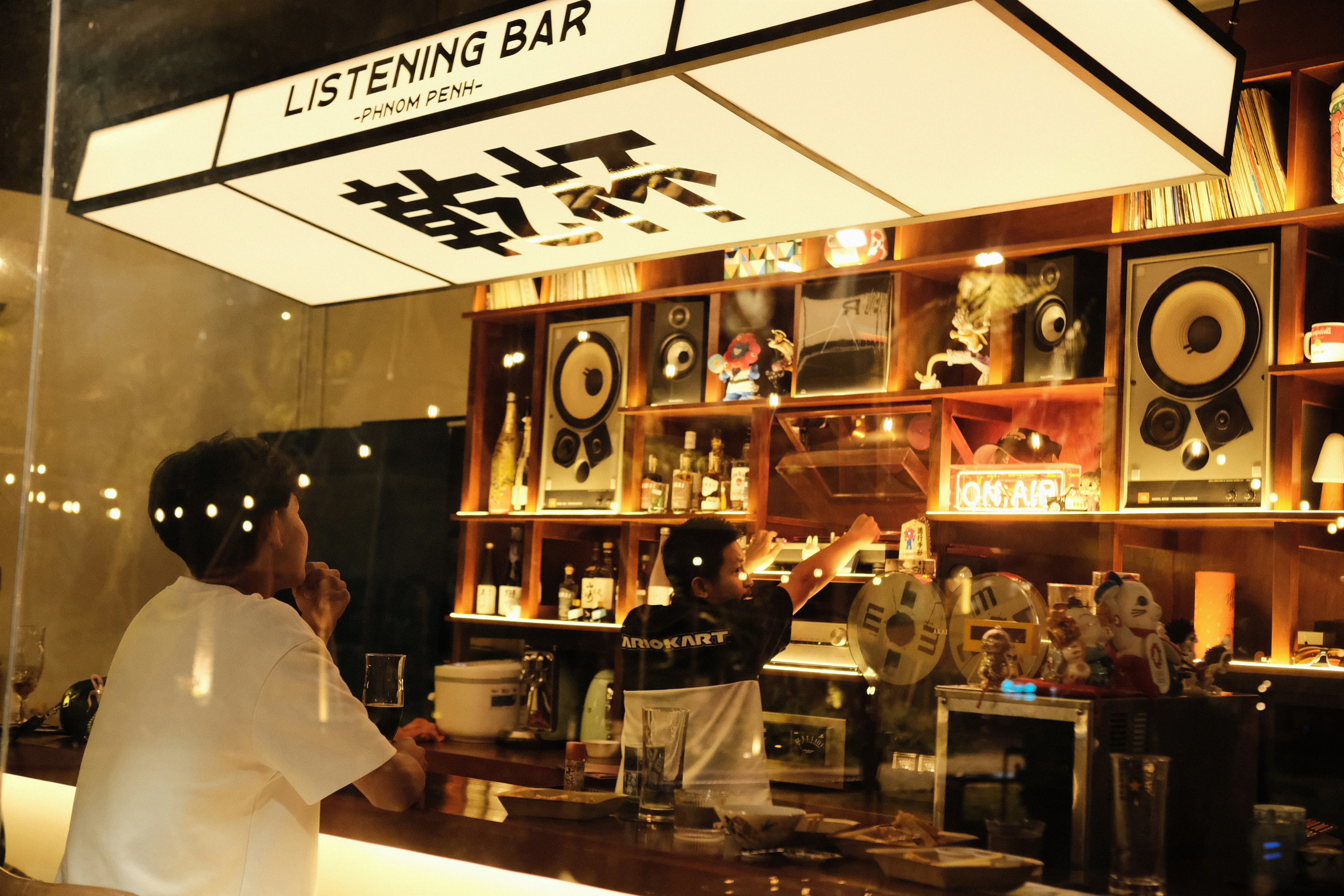 A cozy listening bar scene featuring a bartender engaging with a customer, surrounded by vintage audio equipment and a warm ambiance.