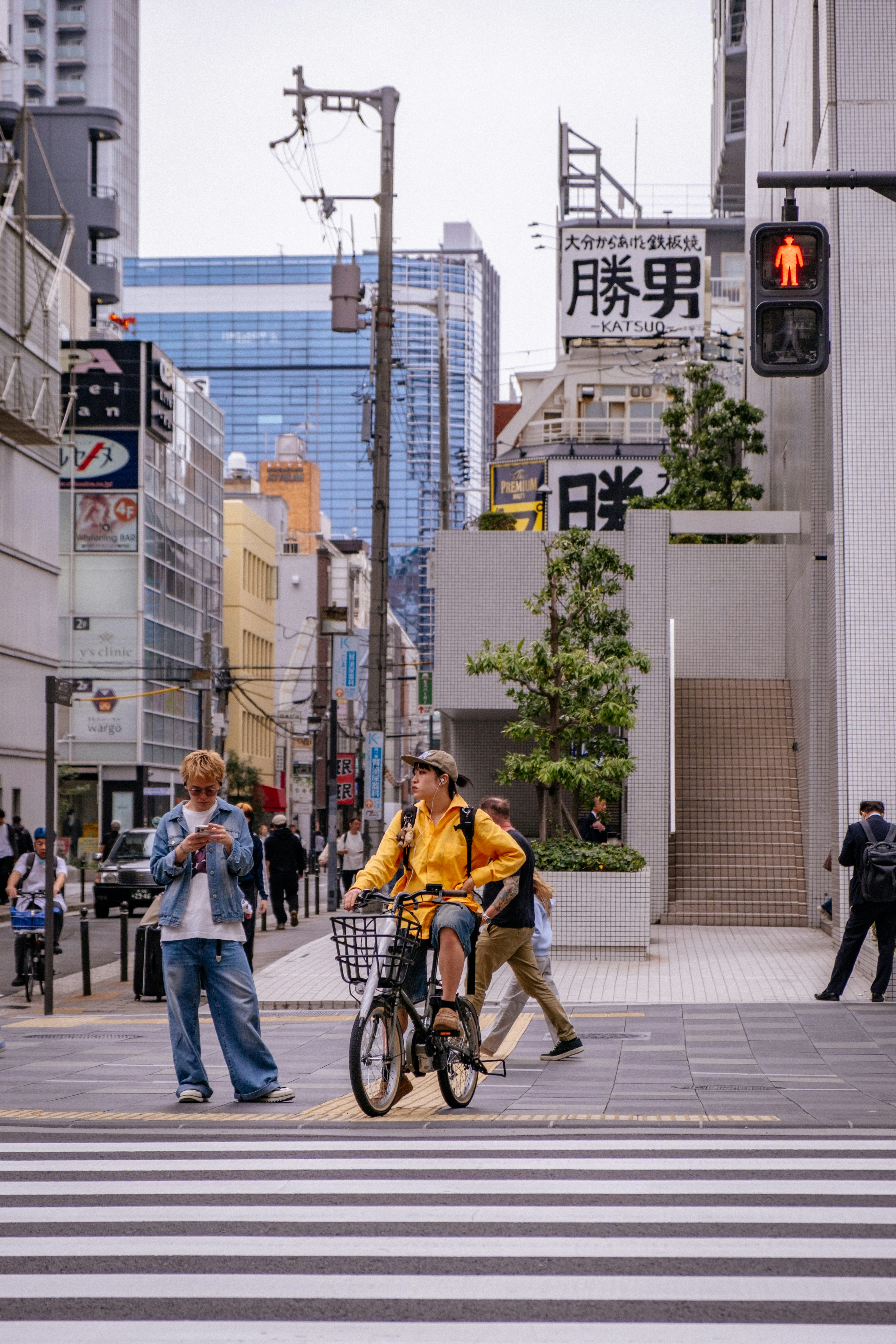 A cyclist pauses at a crosswalk while pedestrians navigate the bustling street, framed by modern architecture and vibrant signage.