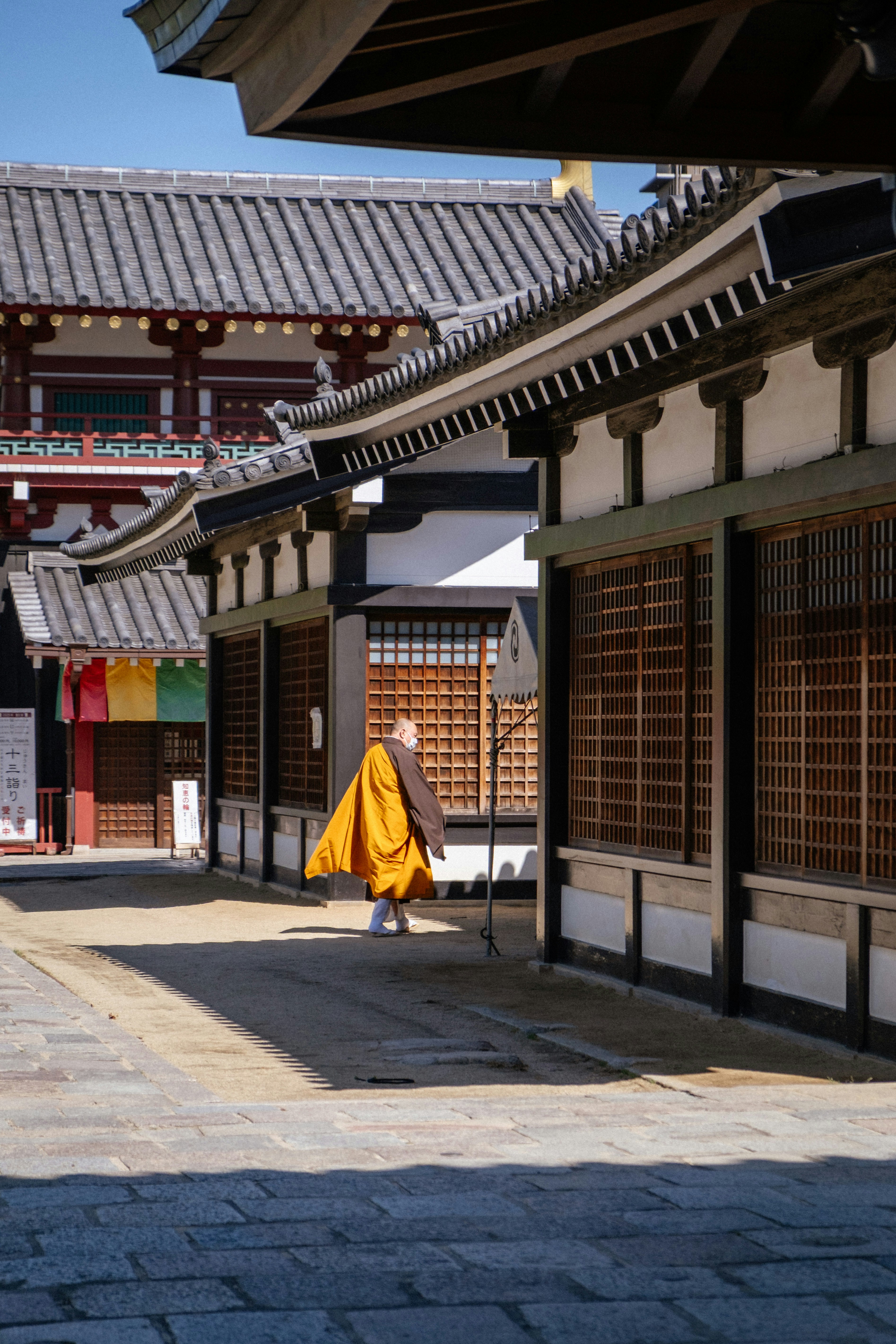Monk in traditional robes walking through a tranquil temple courtyard surrounded by wooden architecture and colorful flags.