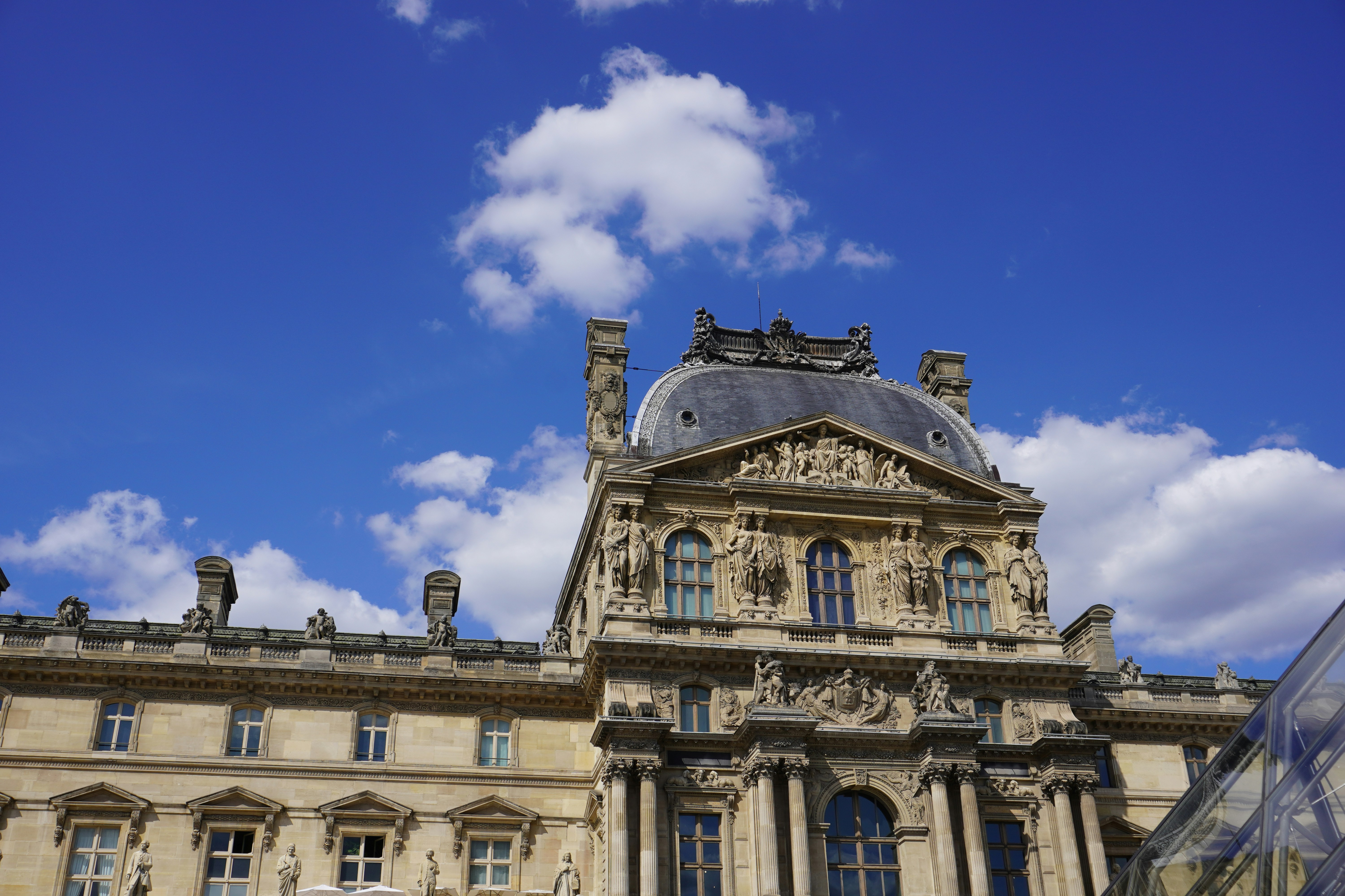 The ornate façade of the Louvre Museum contrasts with a vibrant blue sky dotted with fluffy clouds.