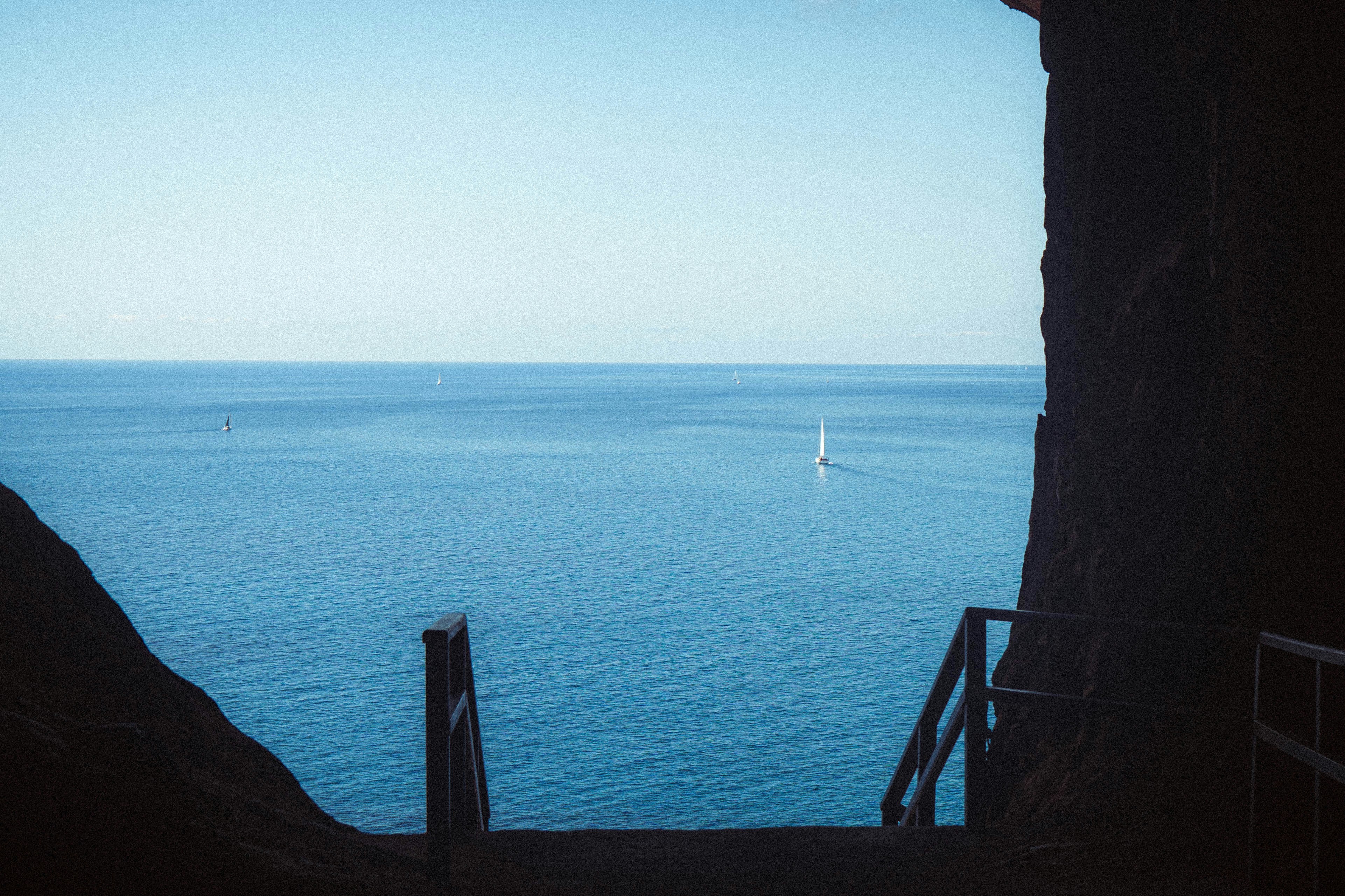 A peaceful view of the open sea from a shadowy cliffside passage, with a lone sailboat drifting across the horizon. Captured during the summer holidays in Mallorca, this moment evokes calm, freedom, and the quiet beauty of the Mediterranean. | A boat sails on a blue sea.