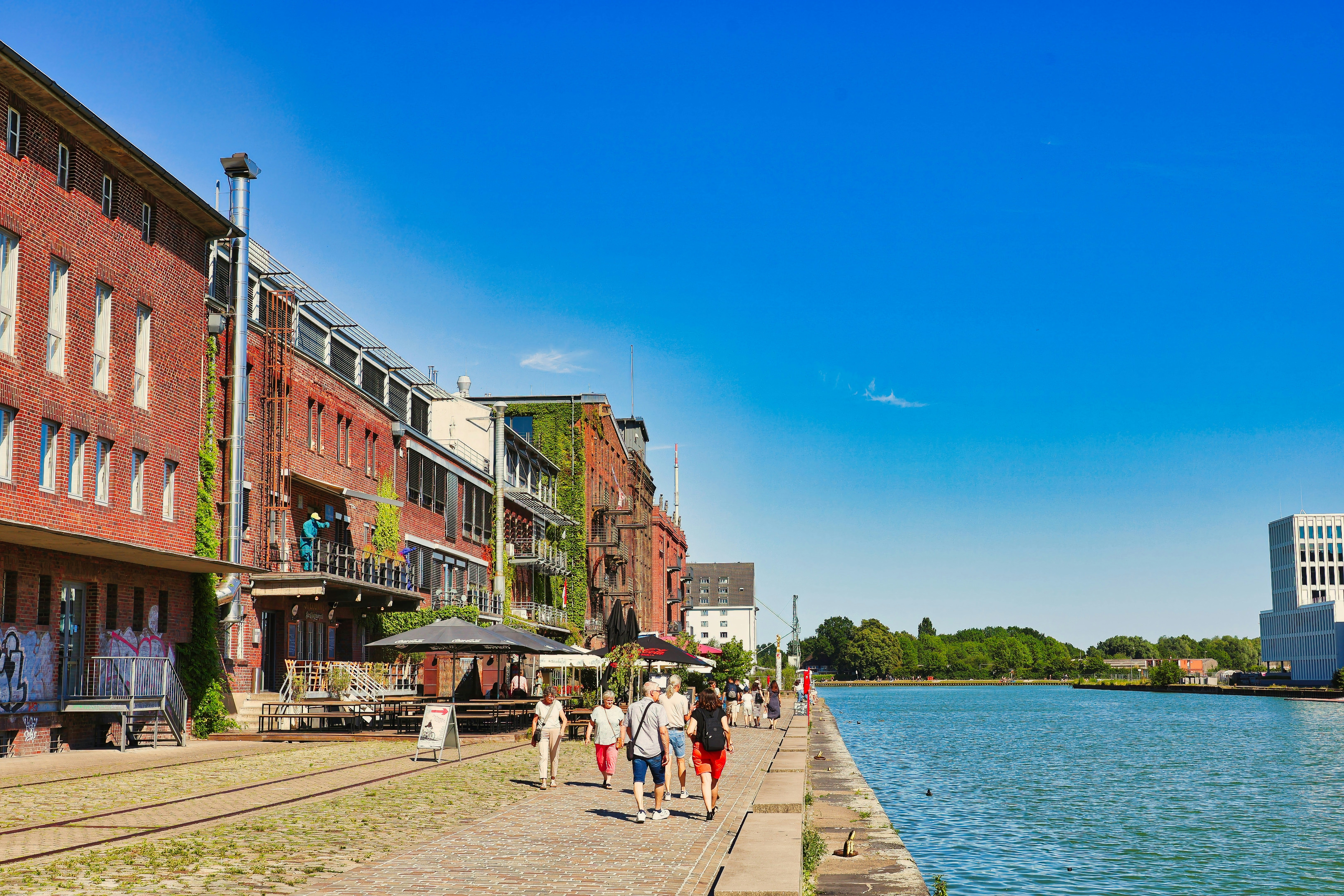 People stroll along a waterfront promenade lined with historic brick buildings and greenery, reflecting a blend of urban life and nature.
