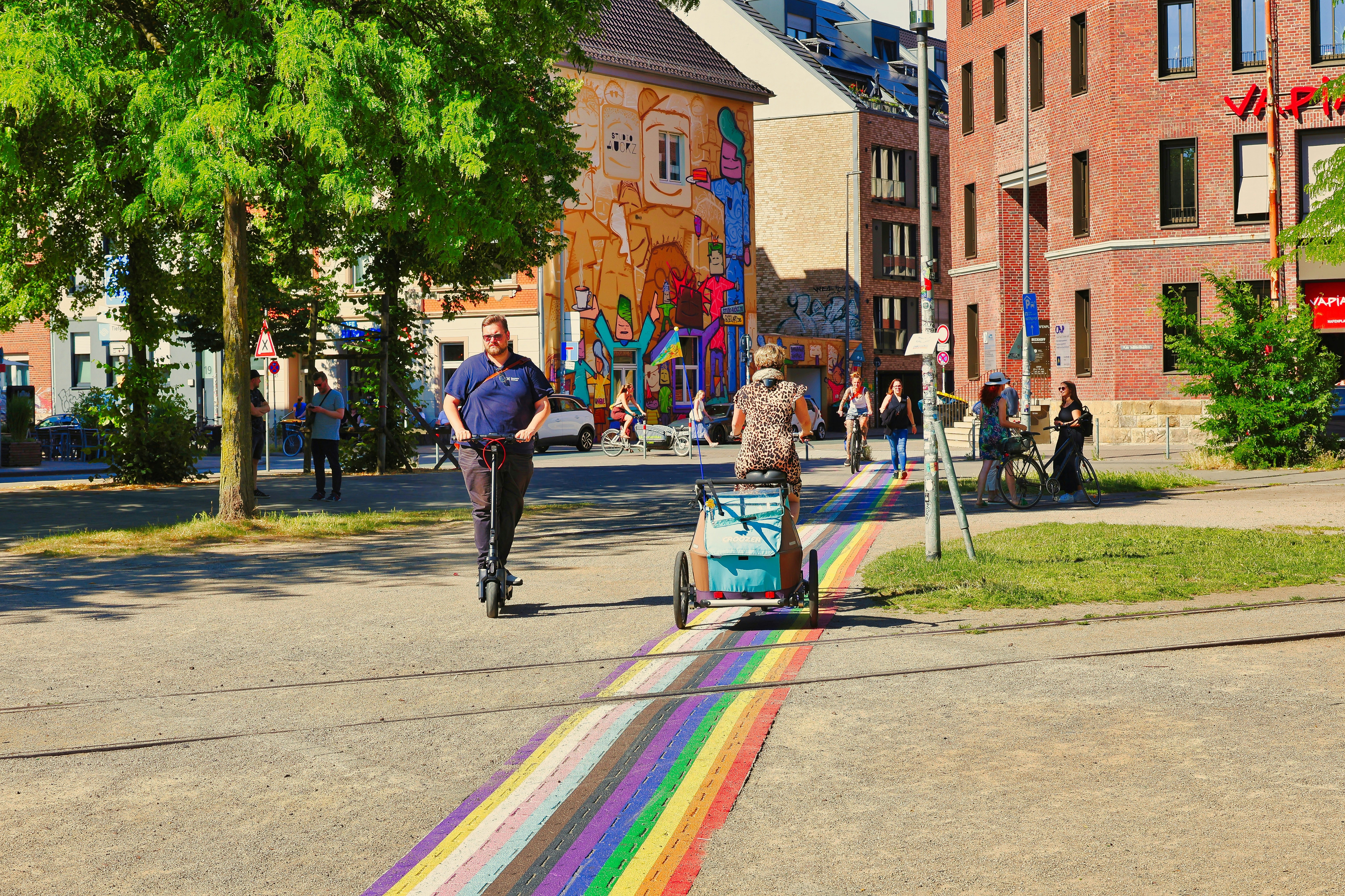 Two individuals traverse a lively street adorned with a rainbow pathway, surrounded by urban art and greenery. The scene captures the essence of community and creativity.