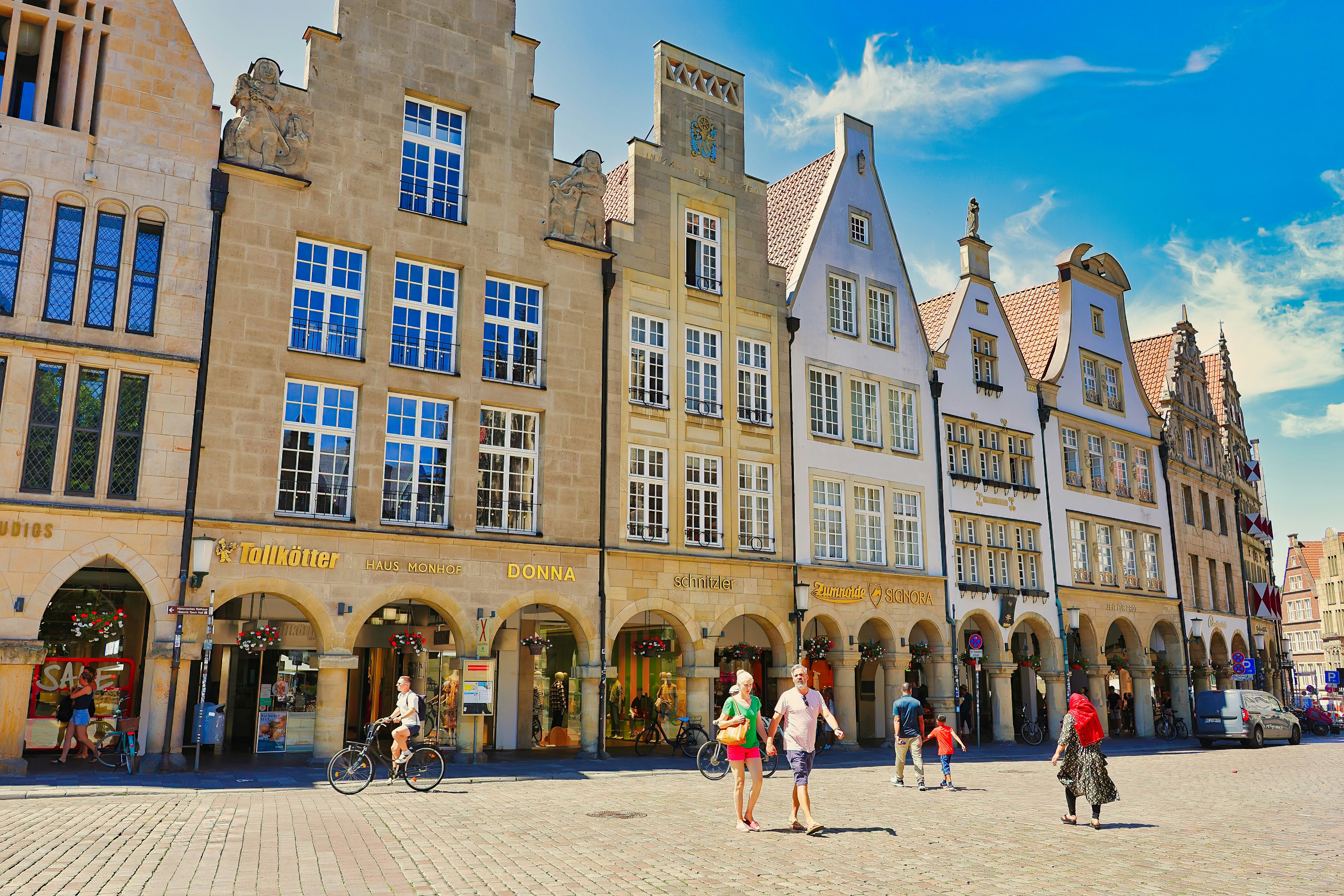 Charming historical buildings line a bustling square, showcasing intricate facades and lively street activity. Pedestrians and cyclists navigate the vibrant scene.