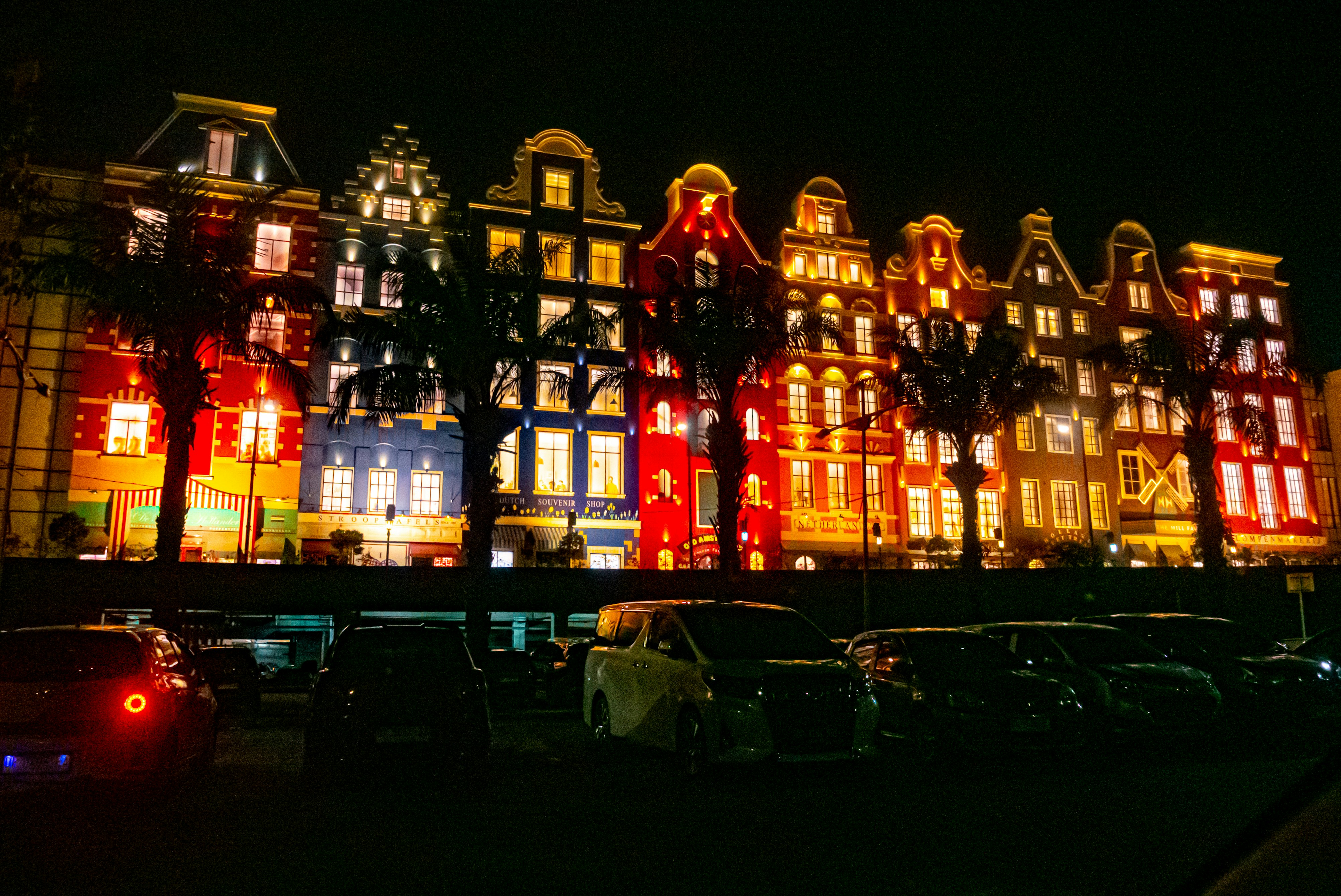 Vibrant Dutch-style buildings illuminated at night, showcasing colorful facades with European architectural charm, framed by silhouettes of palm trees and parked cars in a tropical urban setting. | Illuminated buildings at night with colorful lights.