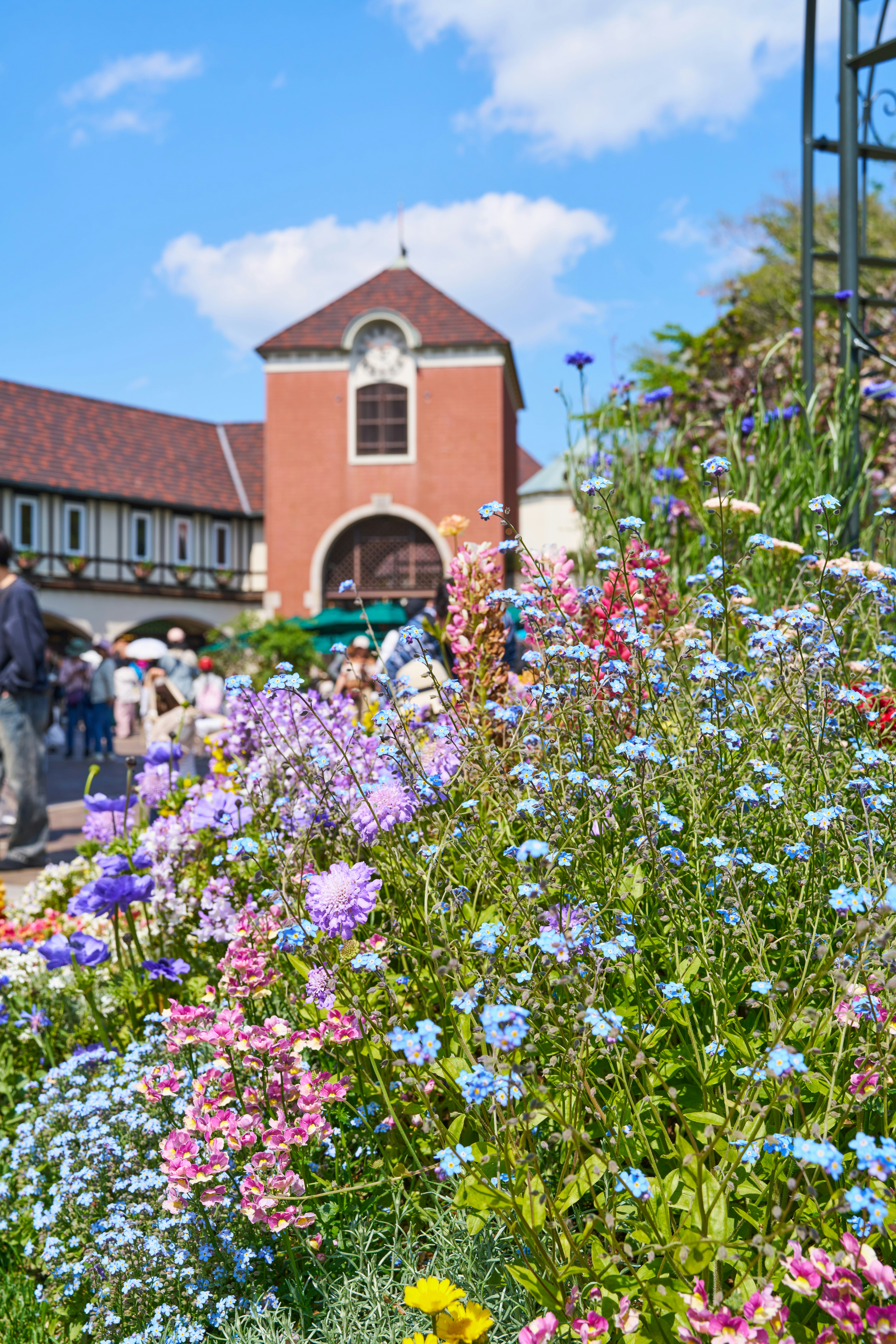 Vibrant flower beds in bloom frame a historic building, bustling with visitors enjoying a sunny day at a market. 