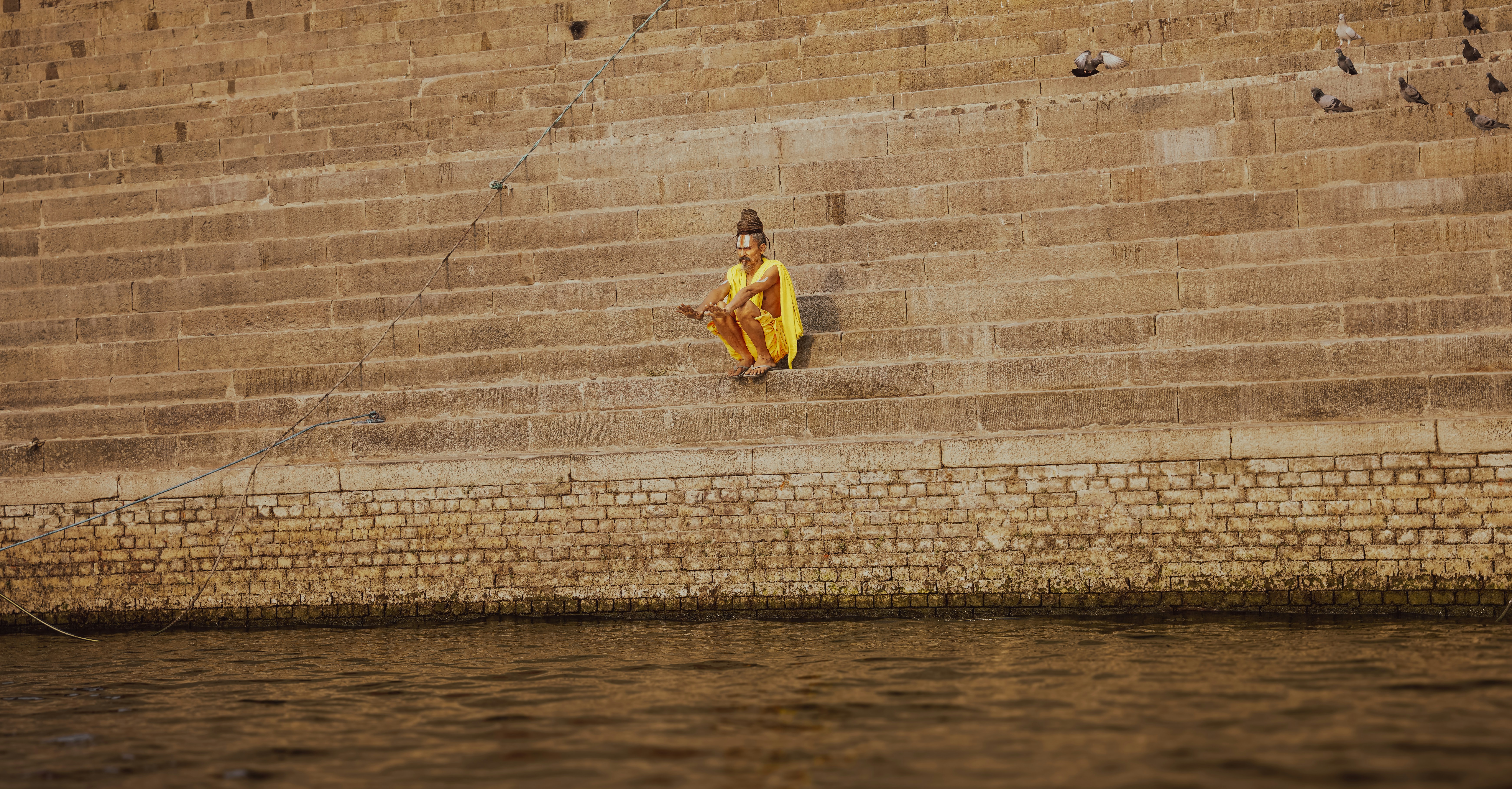 A person in a vibrant yellow outfit sits contemplatively on stone steps by a serene body of water, surrounded by a rustic wall and scattered pigeons above.