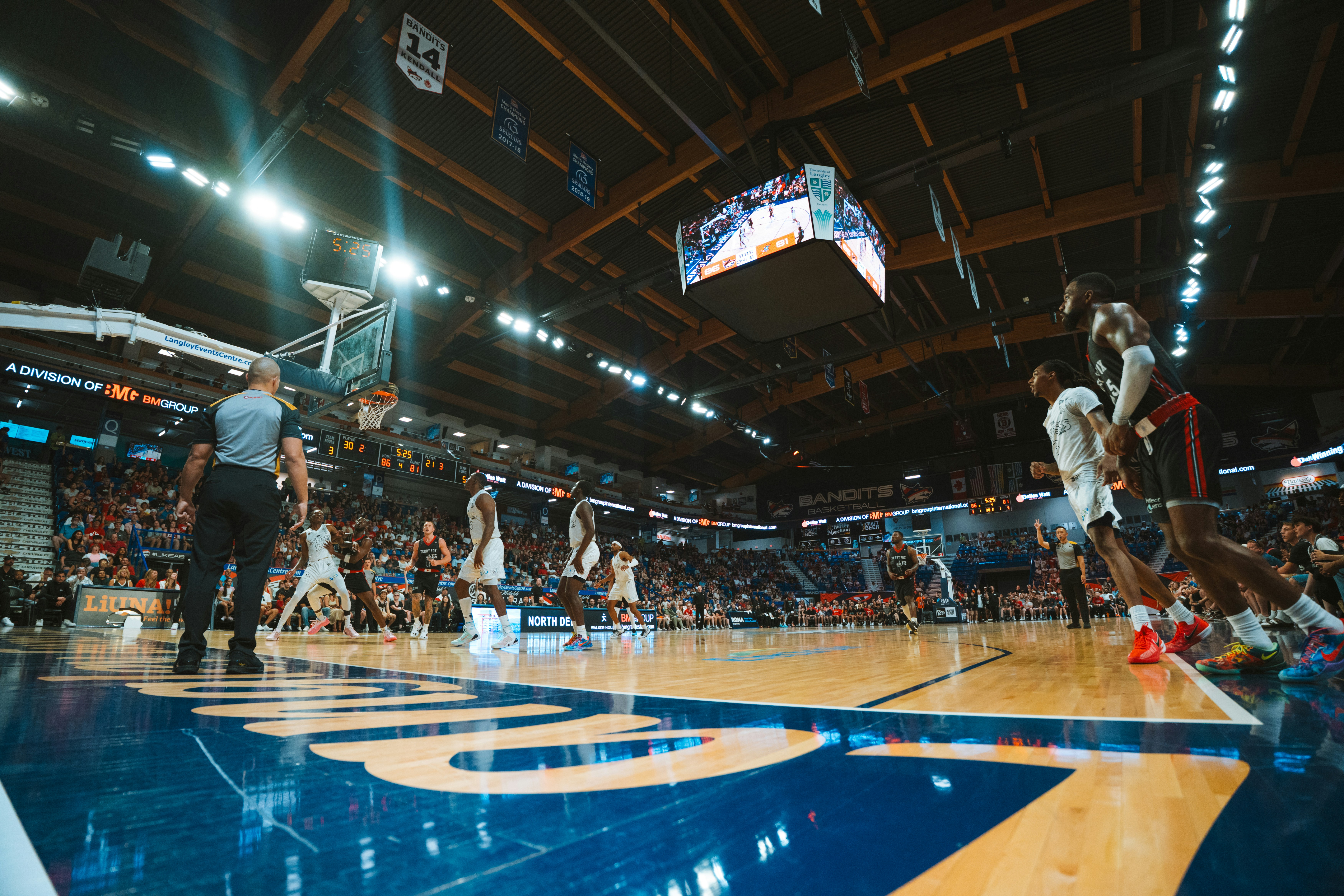 Basketball players in action during a game, with a referee observing from the sidelines. The arena is filled with spectators, creating an energetic atmosphere.