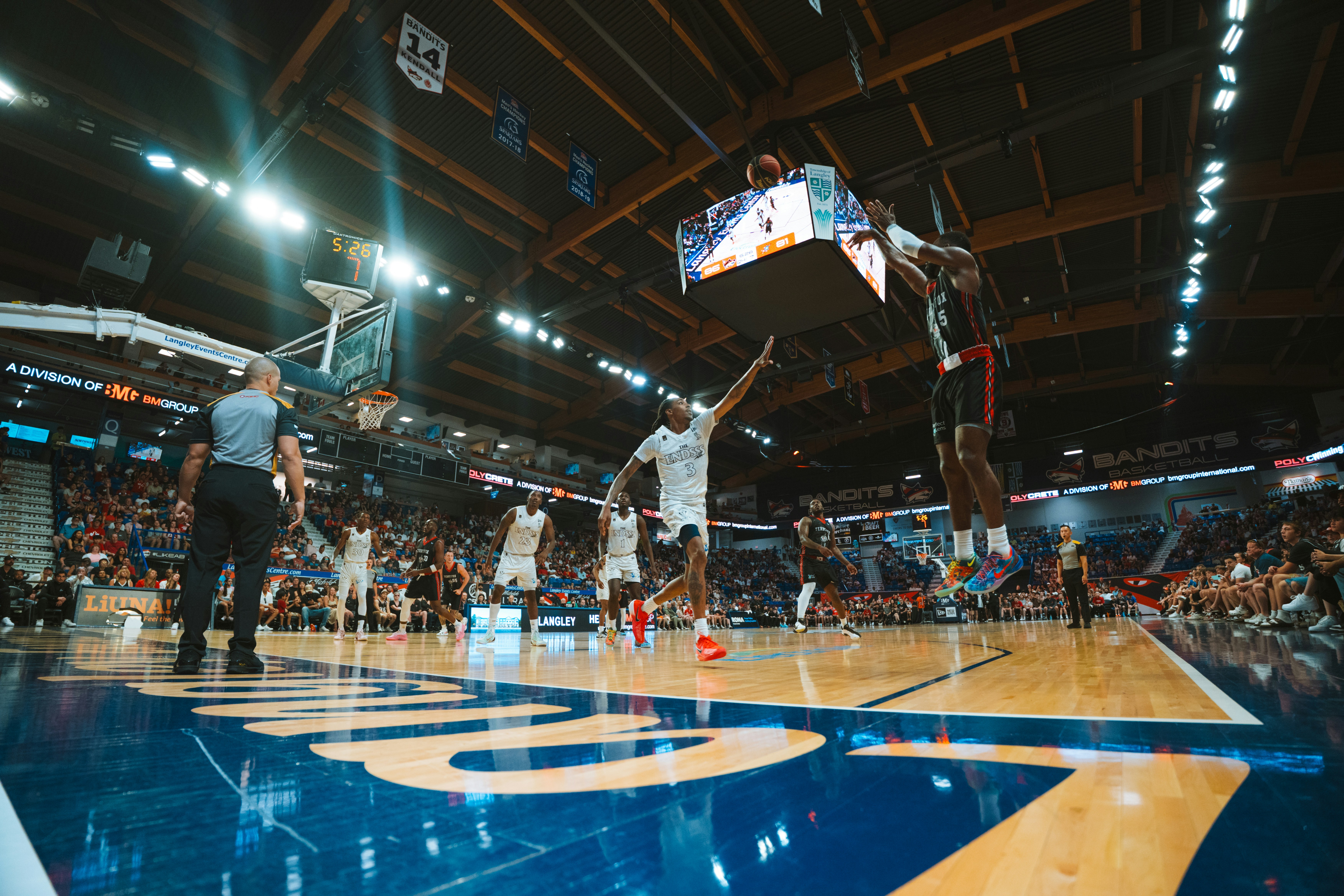 Dynamic basketball scene capturing players in mid-action as one attempts a shot while another reaches for a block. The arena atmosphere is palpable.