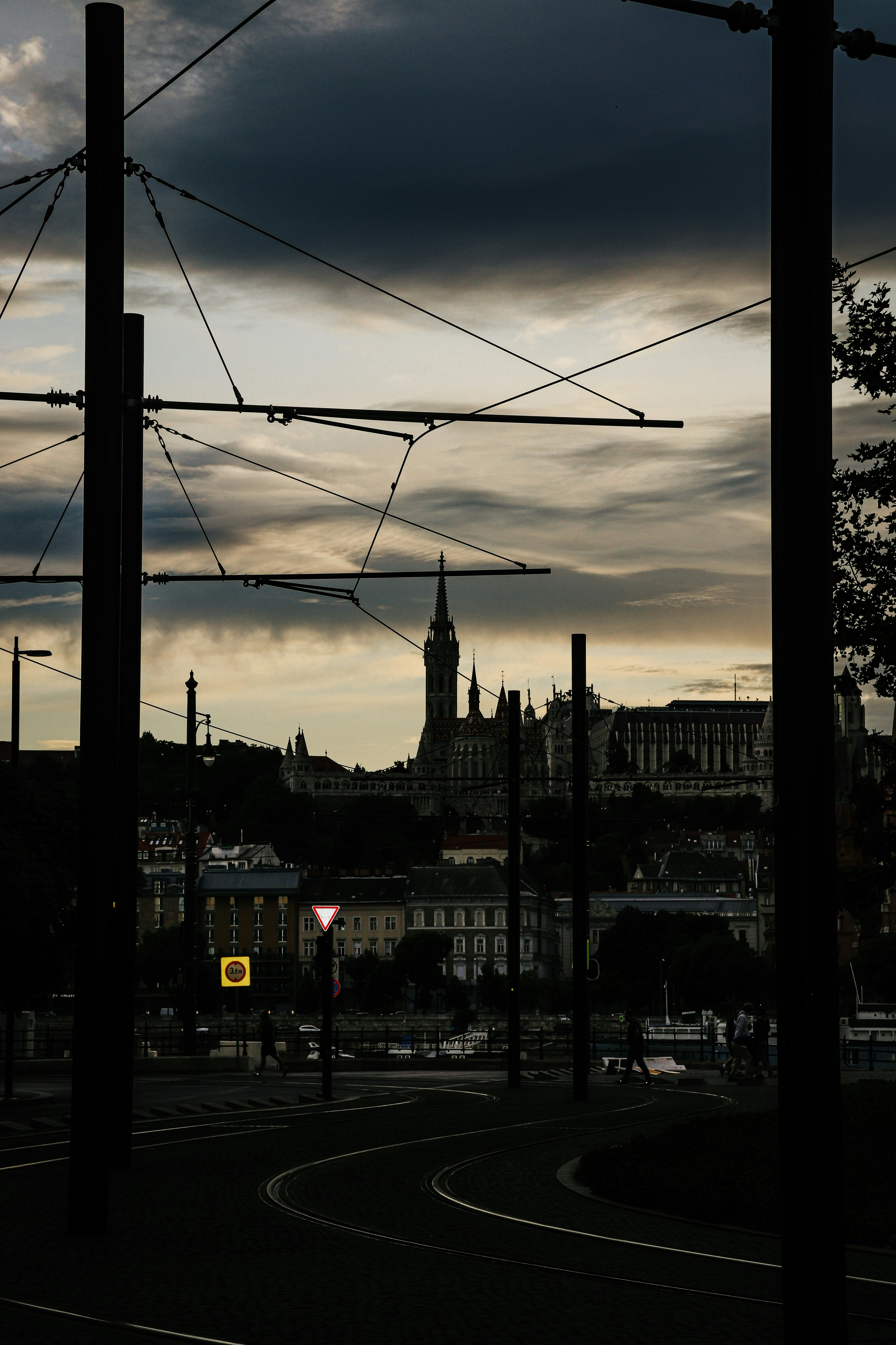 Silhouette of a historic city skyline against a moody sky, framed by tram wires and poles. The scene conveys a sense of urban tranquility as day transitions to night.