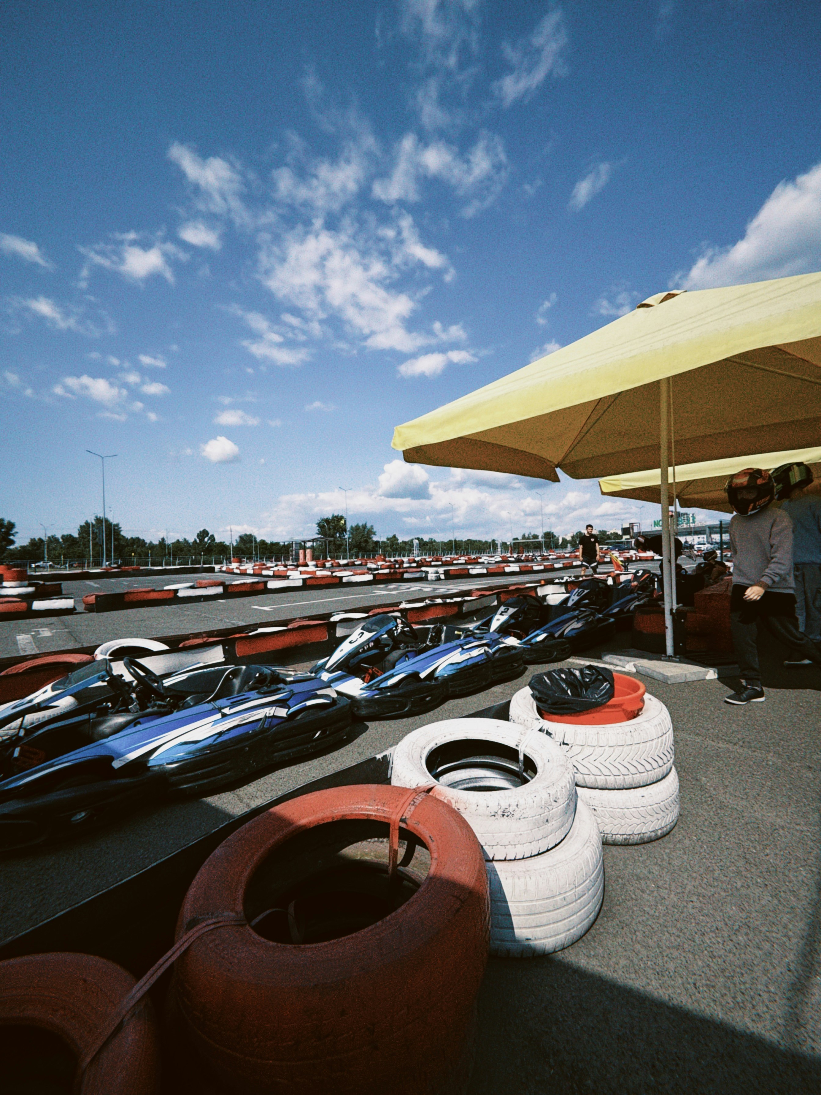 Go-karts lined up under a yellow canopy at a karting track, with vibrant red and white tires in the foreground. A clear blue sky adds to the excitement of the scene.