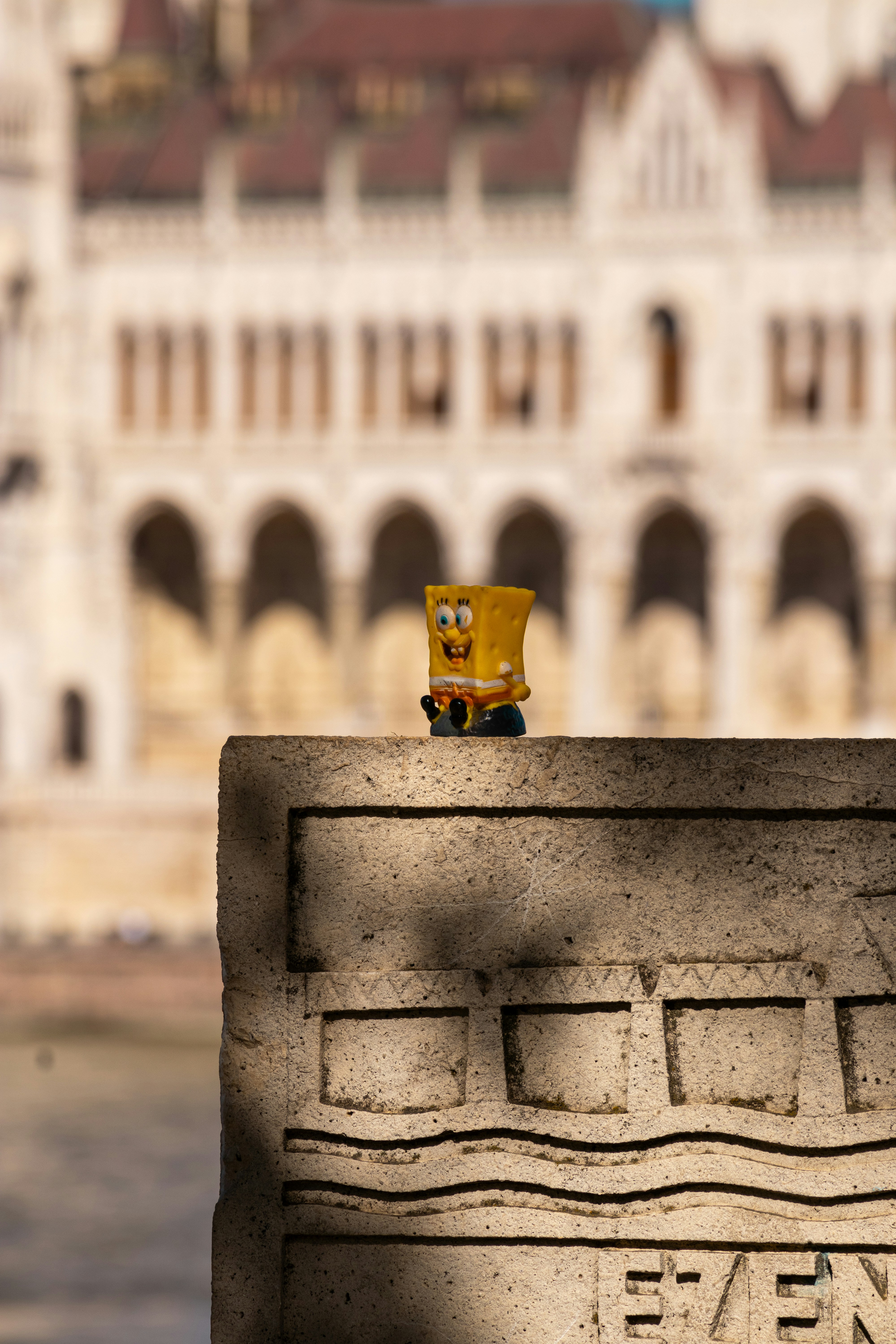 SpongeBob SquarePants figurine perched on a stone ledge with a blurred historic building in the background. The playful contrast between the toy and the architecture adds charm.