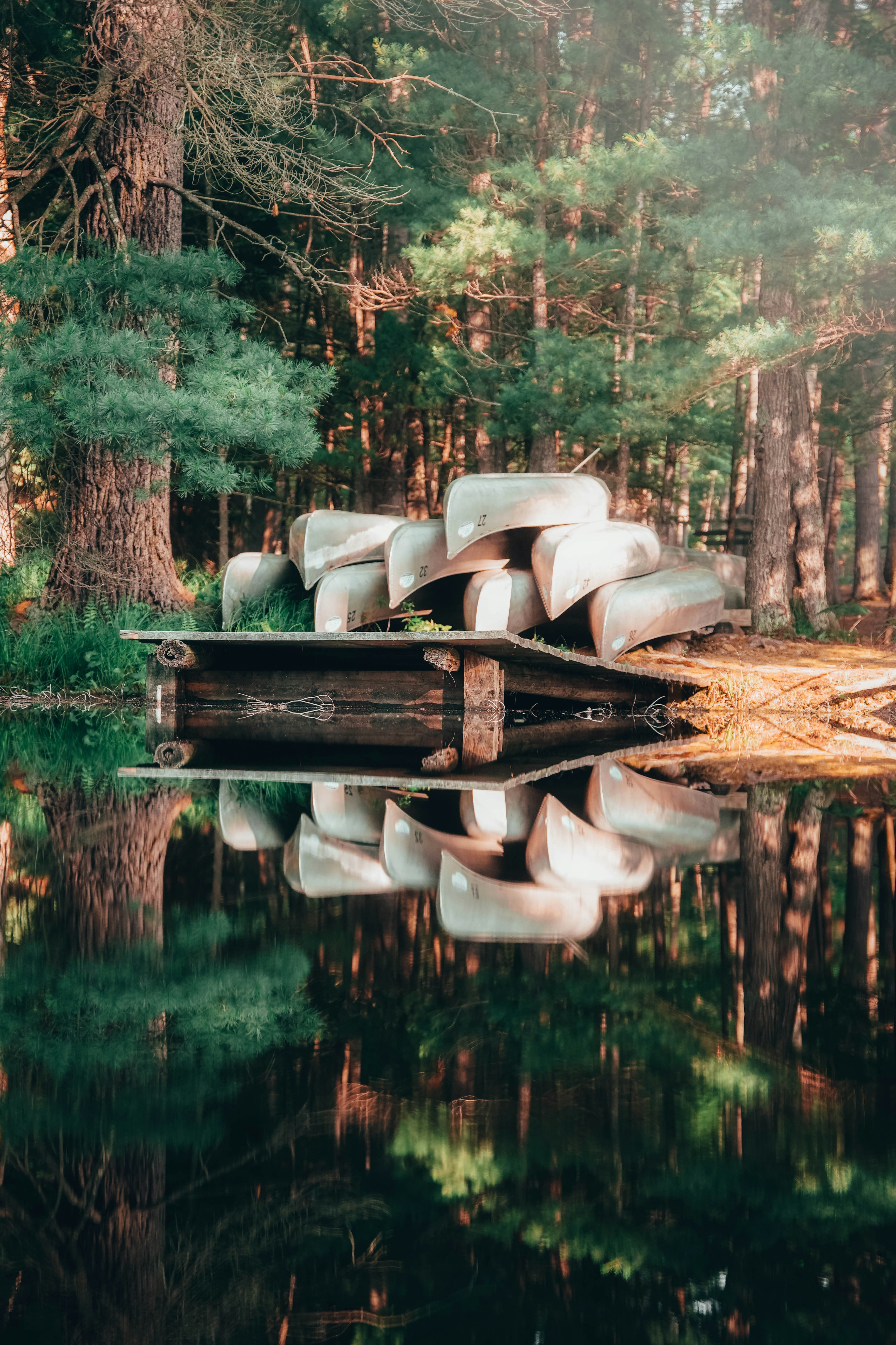 Canoes stacked neatly on a dock, with their colorful reflections shining in the calm water below. | Canoes rest on a dock beside a tranquil lake.