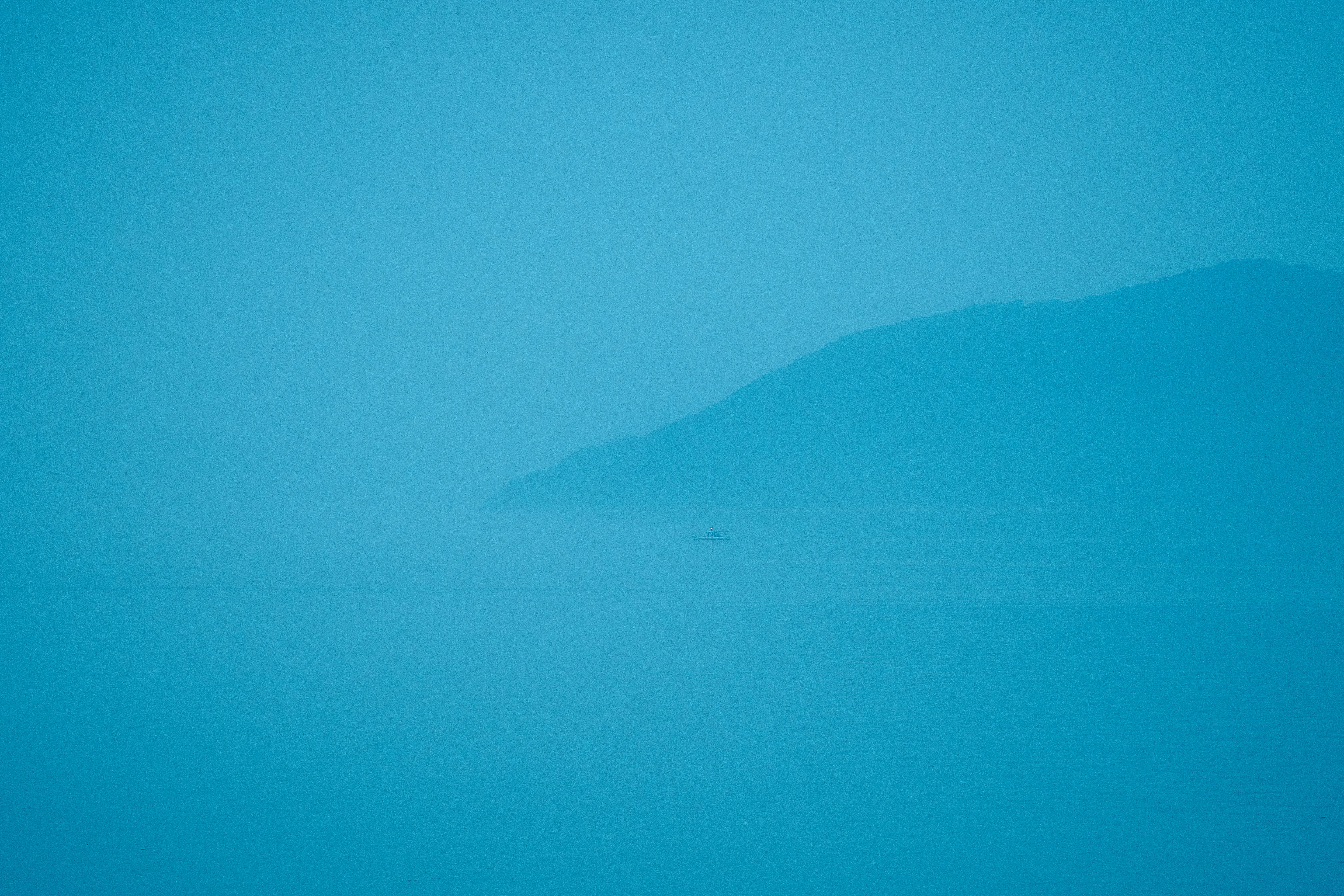 A solitary boat drifts across a serene, mist-covered lake, with distant hills fading into the blue haze.