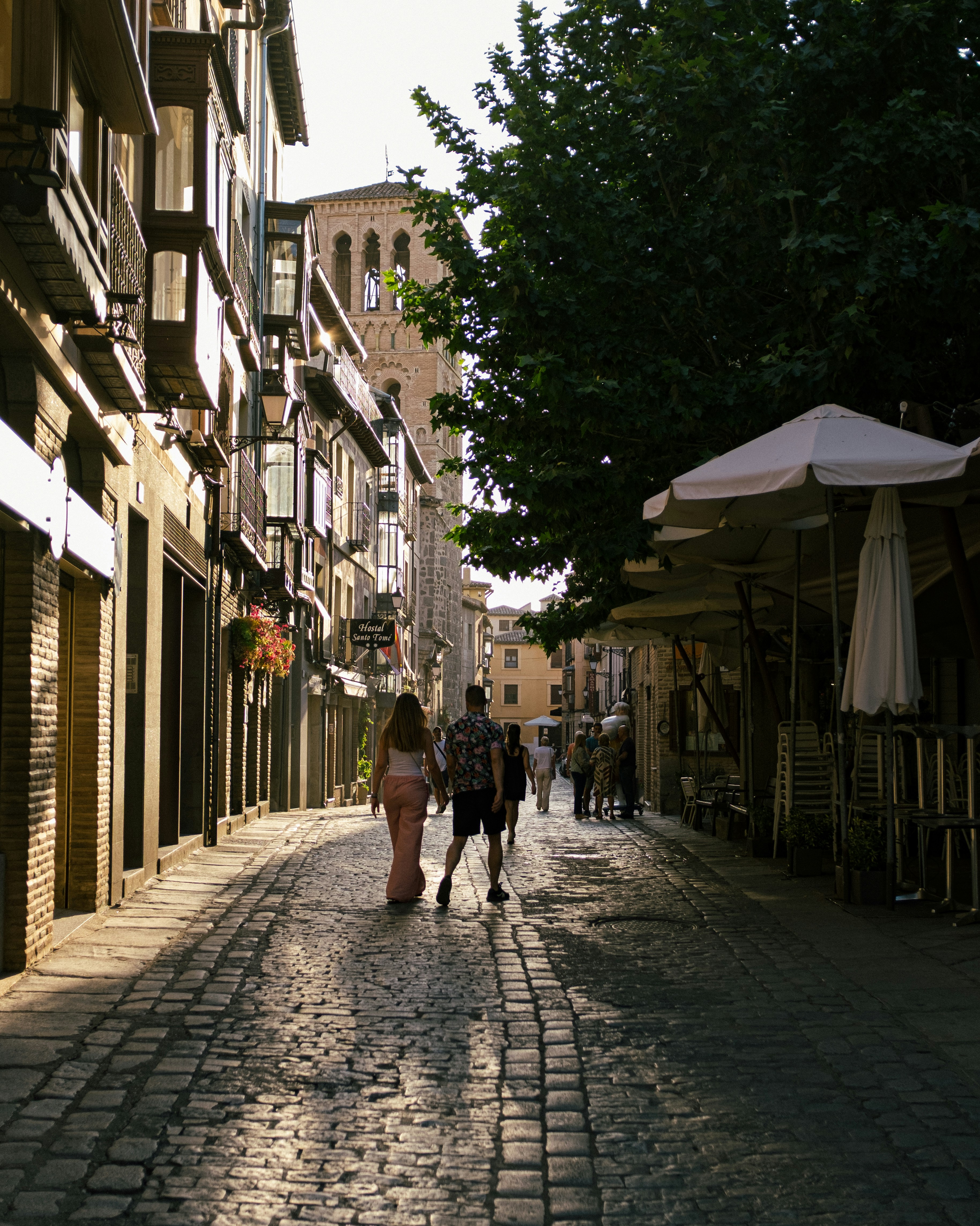 Couple walking hand-in-hand down a sunlit cobblestone street, flanked by historic buildings and vibrant greenery. The scene captures the essence of leisurely urban exploration.